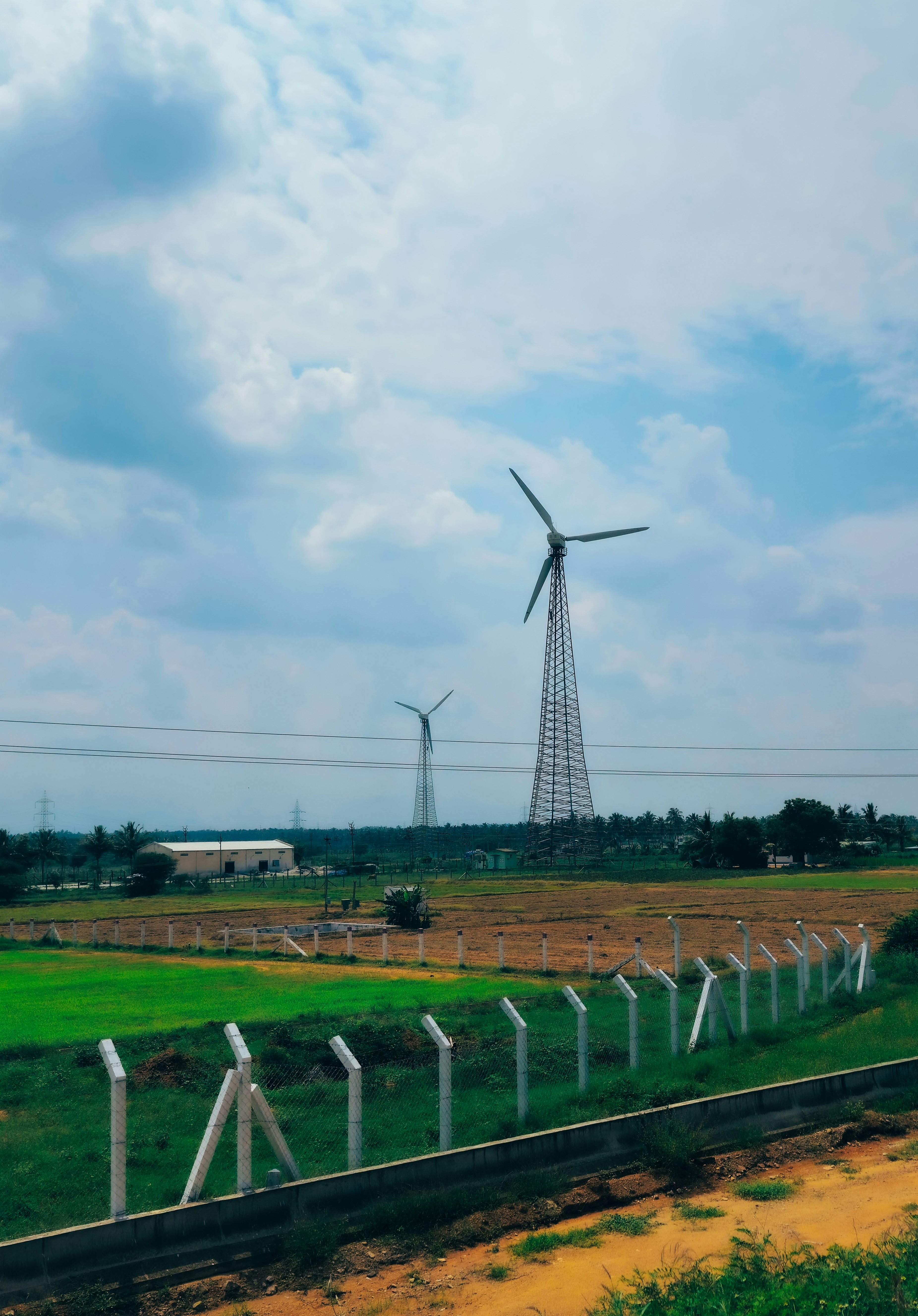 Photograph of a wind turbine dominating the midground above a fence and open fields under a bright blue sky.