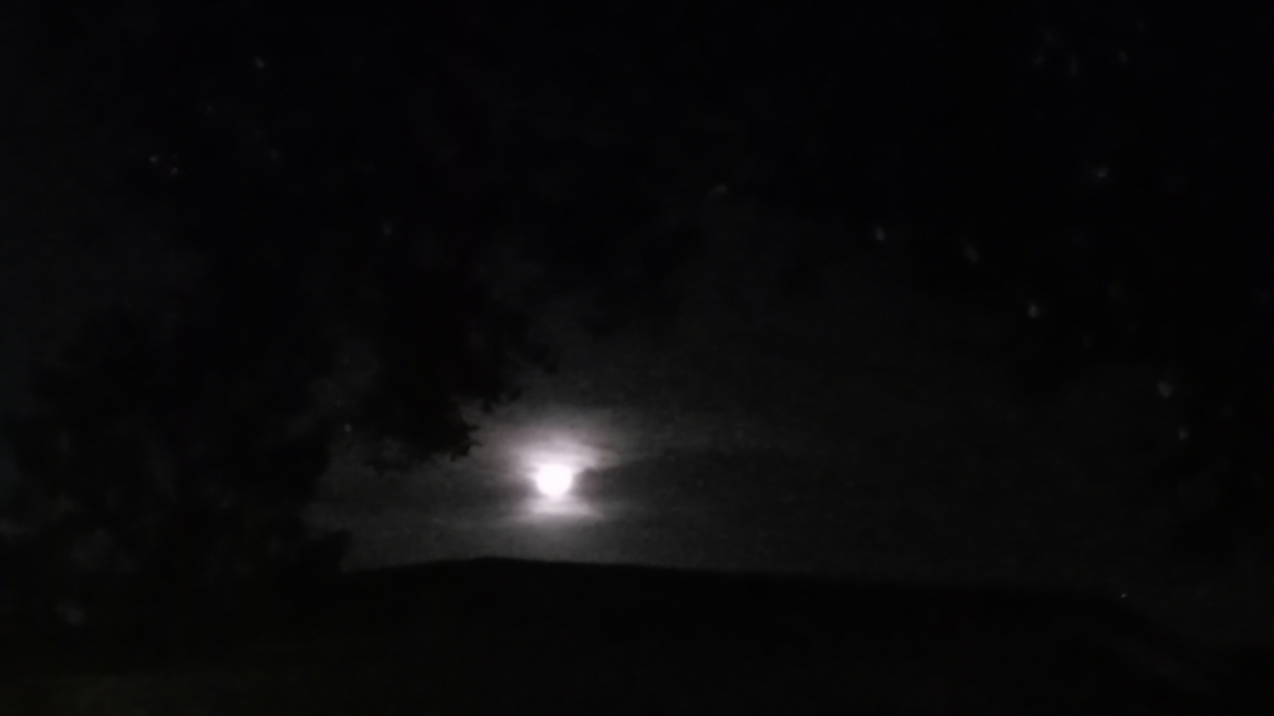 Night photograph of a bright moon peeking through wispy clouds over a dark ridge with silhouetted trees.