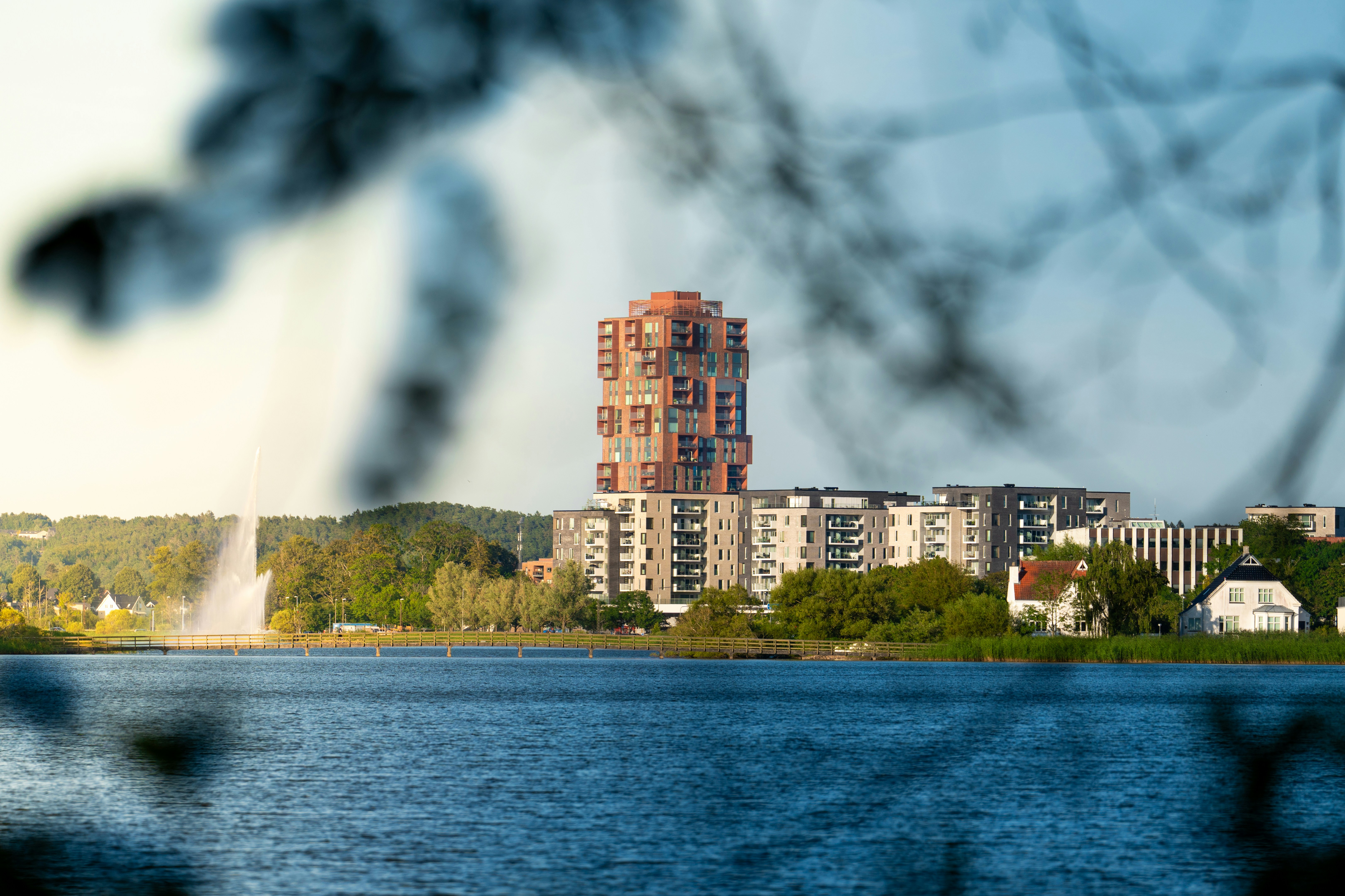 Modern tower rises behind a line of lower buildings across a calm lake, framed by soft-focus branches.