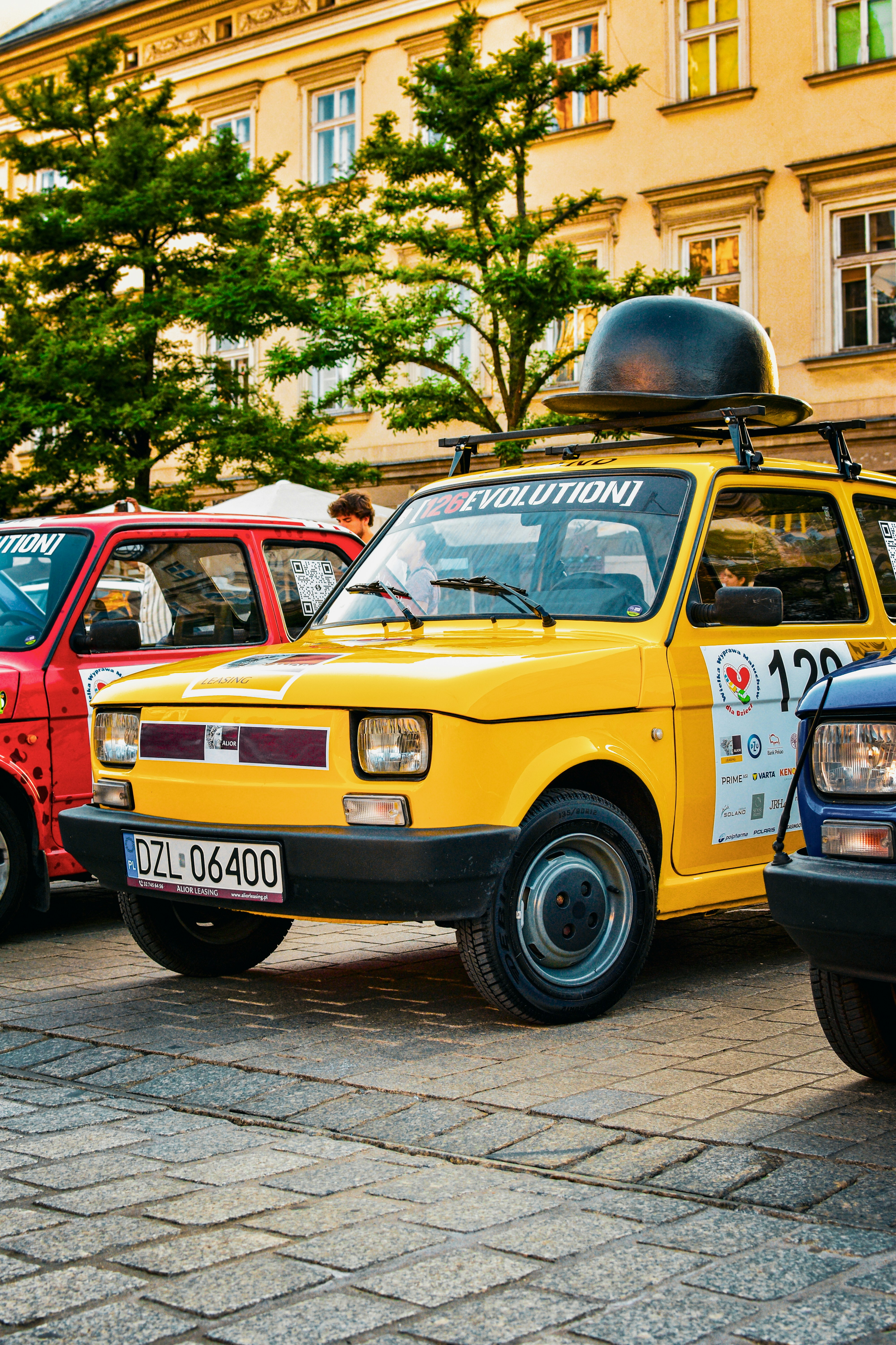 Yellow taxi with rooftop beacon anchors a row of vintage cars in a sunlit city plaza