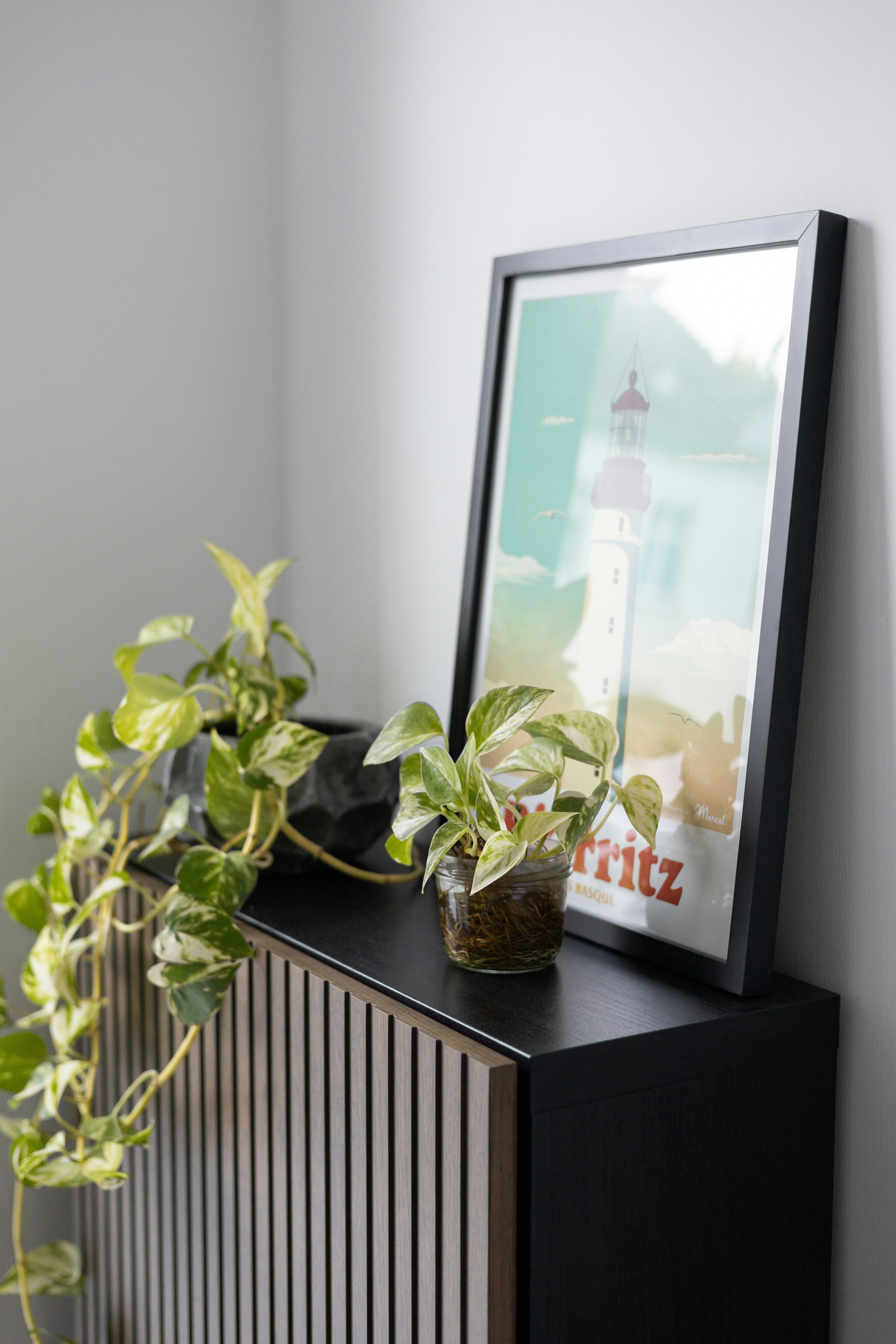 A minimalist home decor scene with a framed poster, a potted plant, and a black wooden shelf.