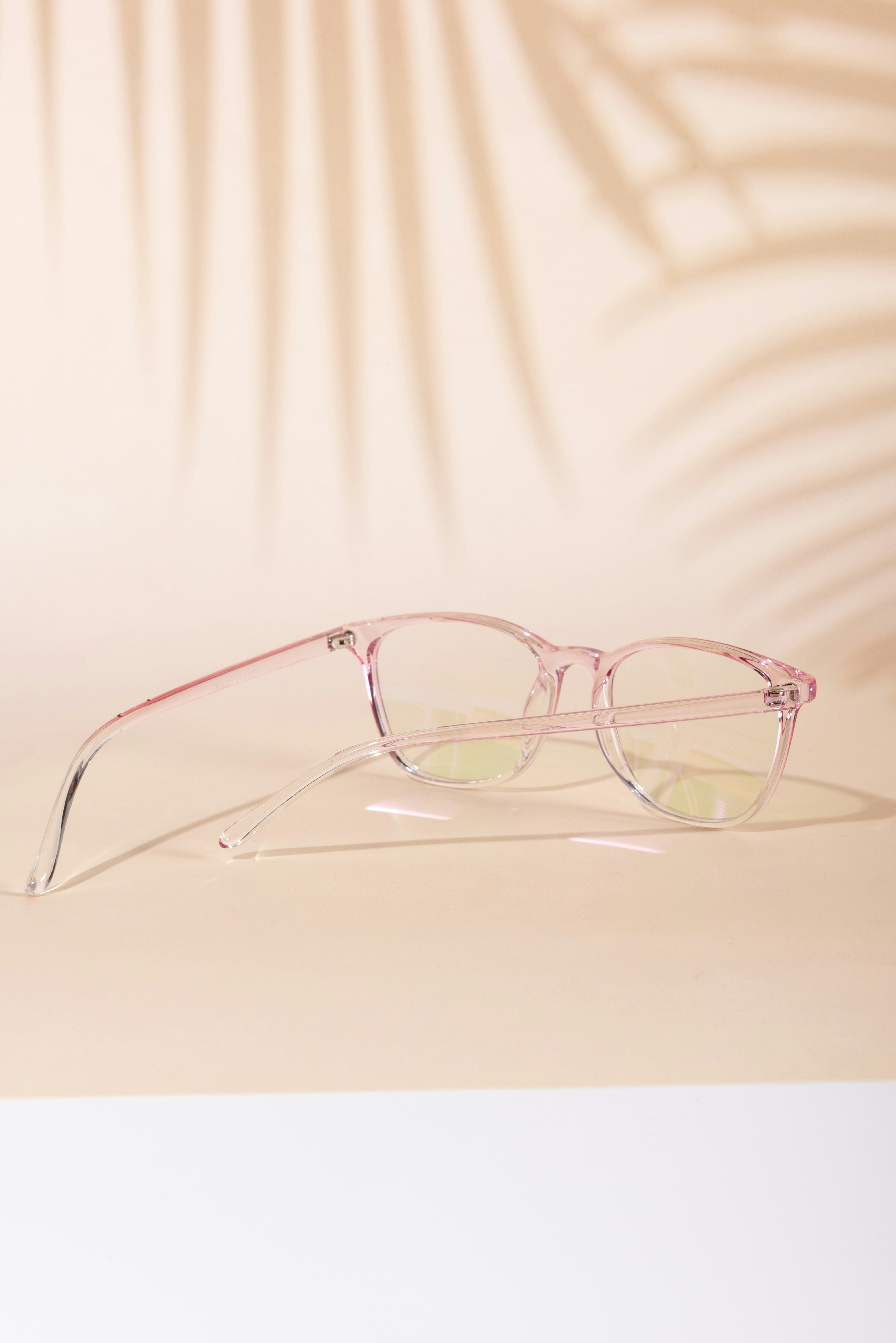 Pink-tinted eyeglasses resting on a pale surface with soft palm shadows
