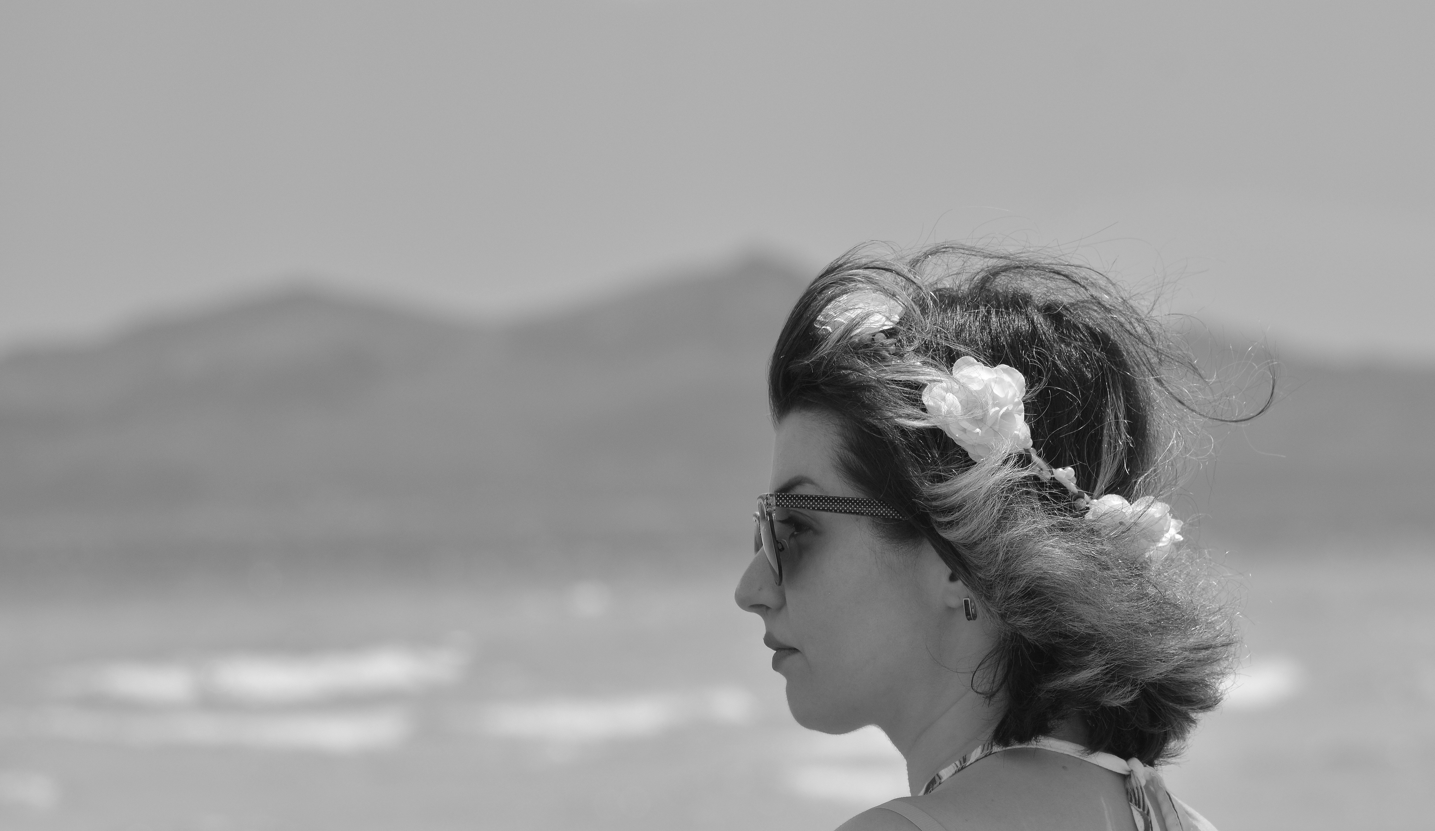 Woman with floral hair accessory gazes towards distant mountains at the beach.
