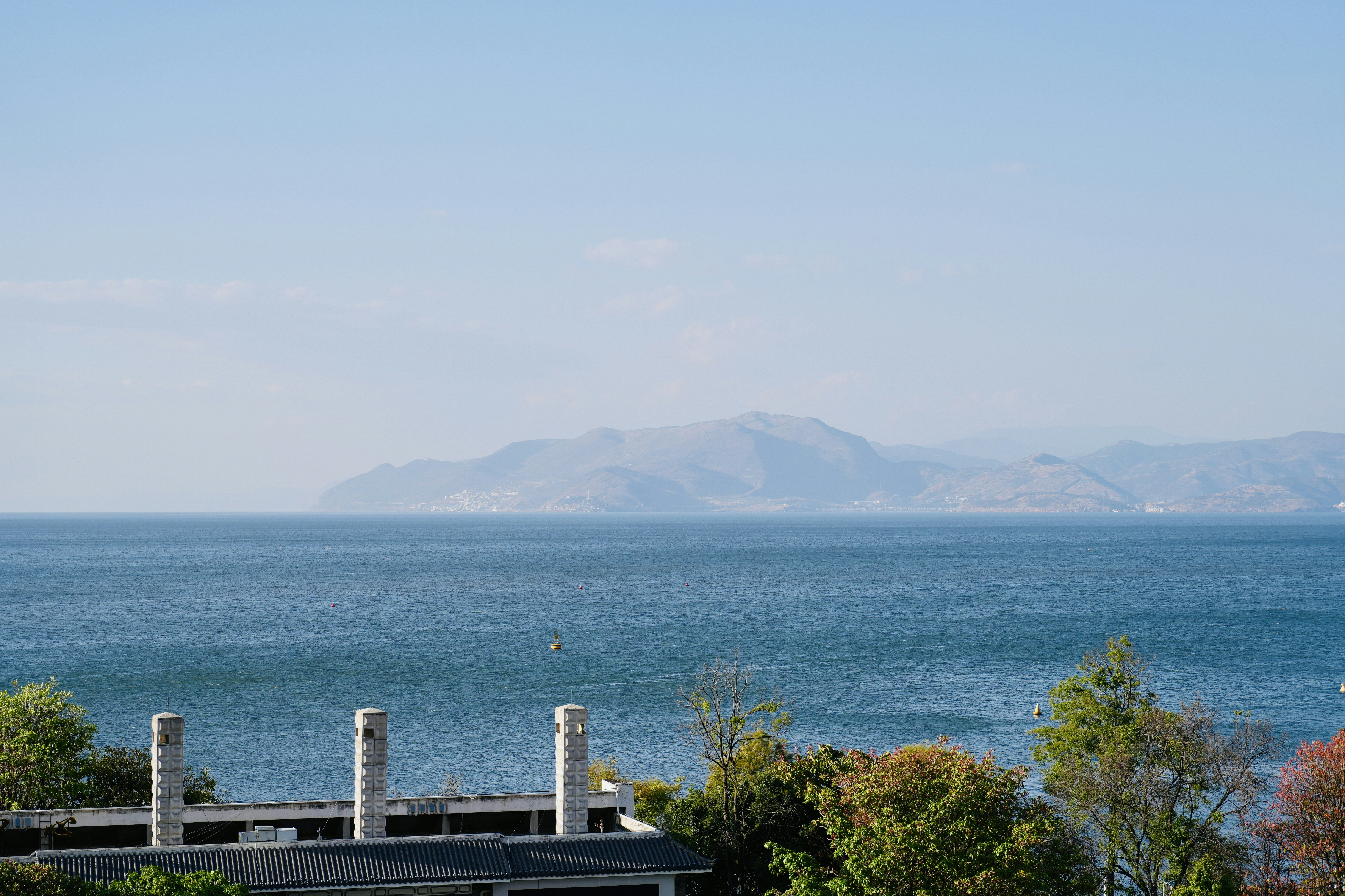 Distant island viewed across calm blue sea with greenery and buildings in the foreground.