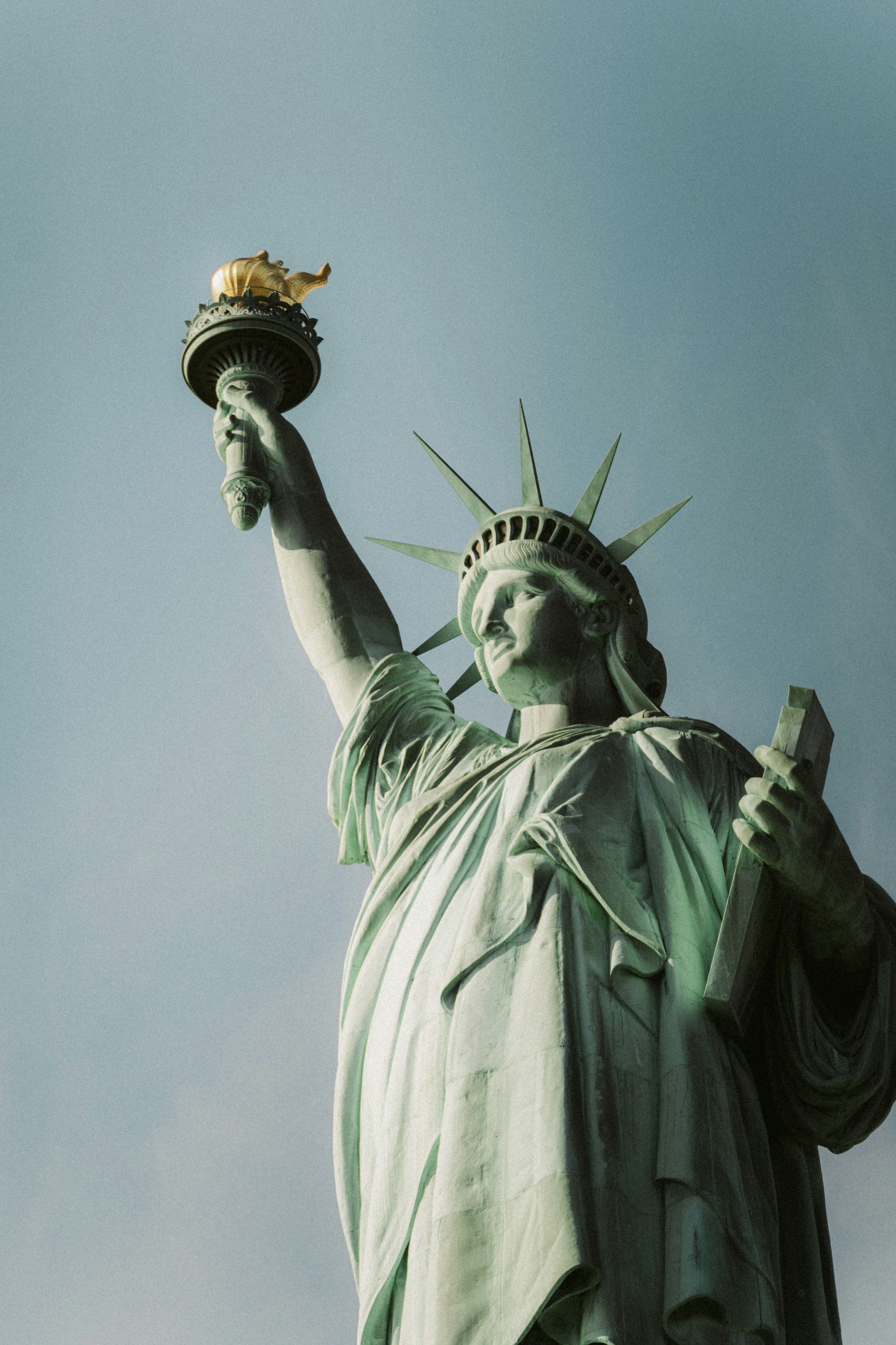 Statue of Liberty with detailed robe folds and crown spikes against a pale blue sky.