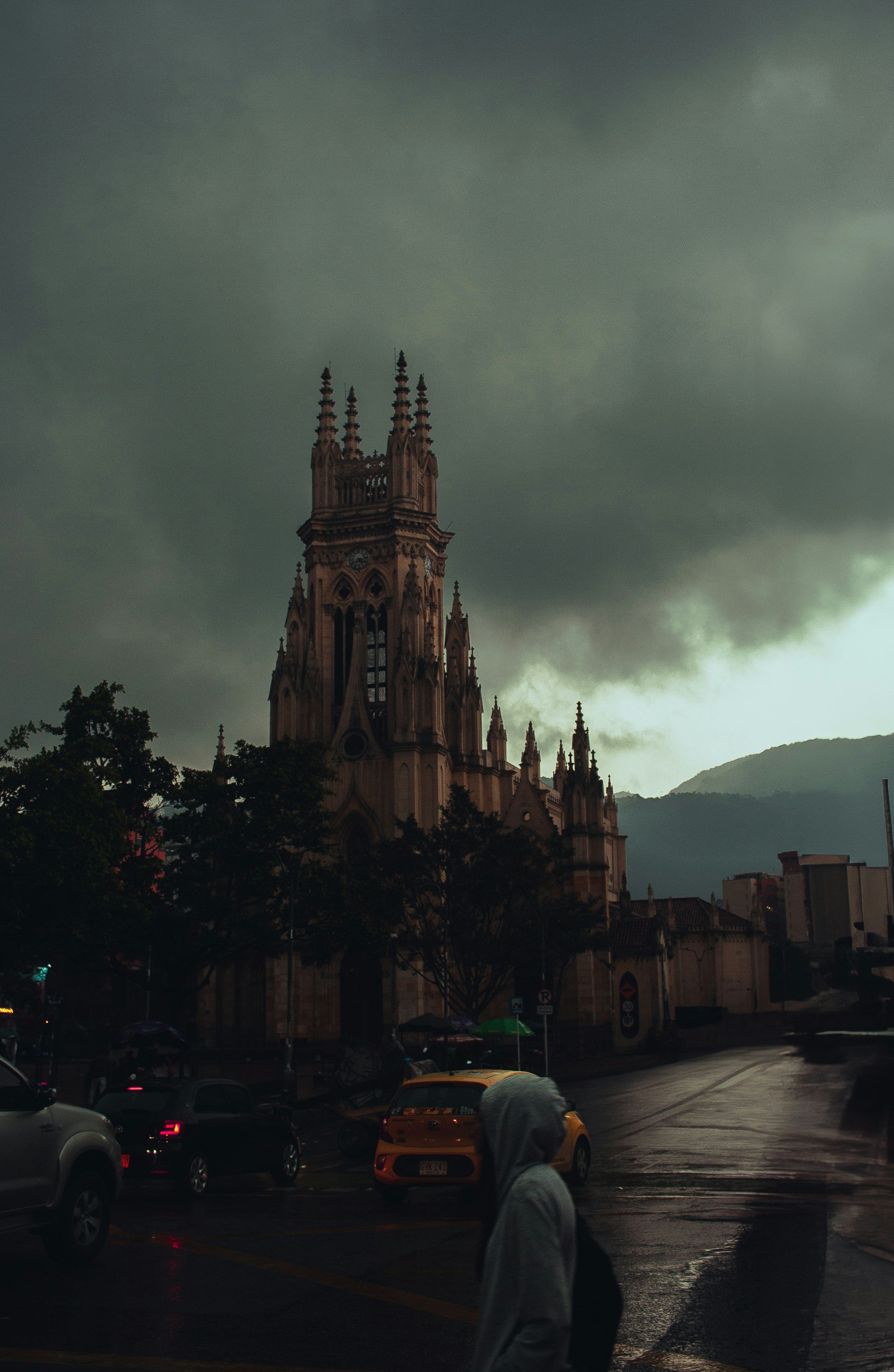 Rain-soaked street with a towering Gothic cathedral piercing stormy skies. A hooded figure moves in the foreground, adding human scale.