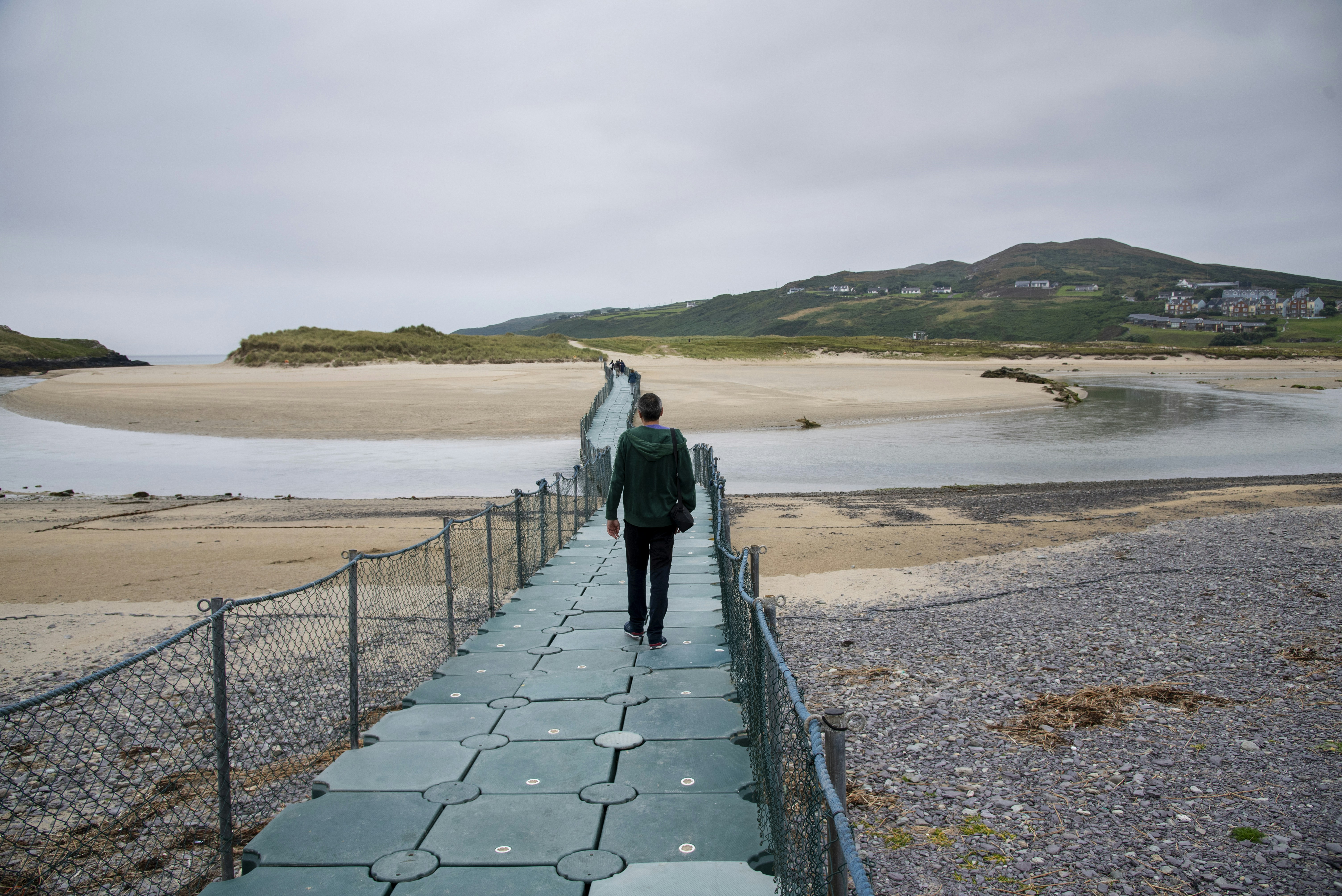 A solitary figure walks along a floating pathway leading to a sandy beach, framed by gentle hills and a cloudy sky.