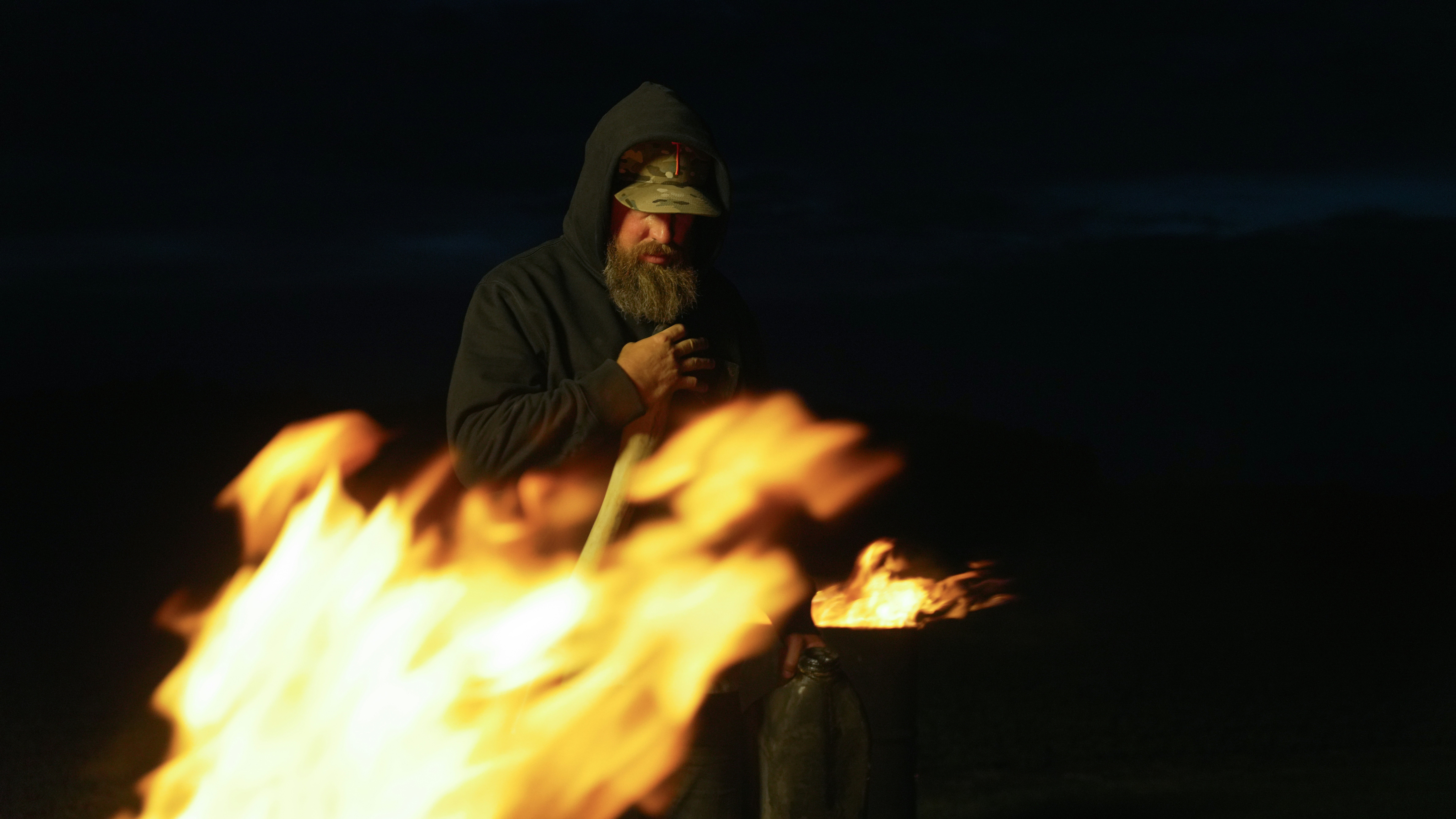 Man in a hoodie stands thoughtfully in front of a blazing fire under a dark sky.