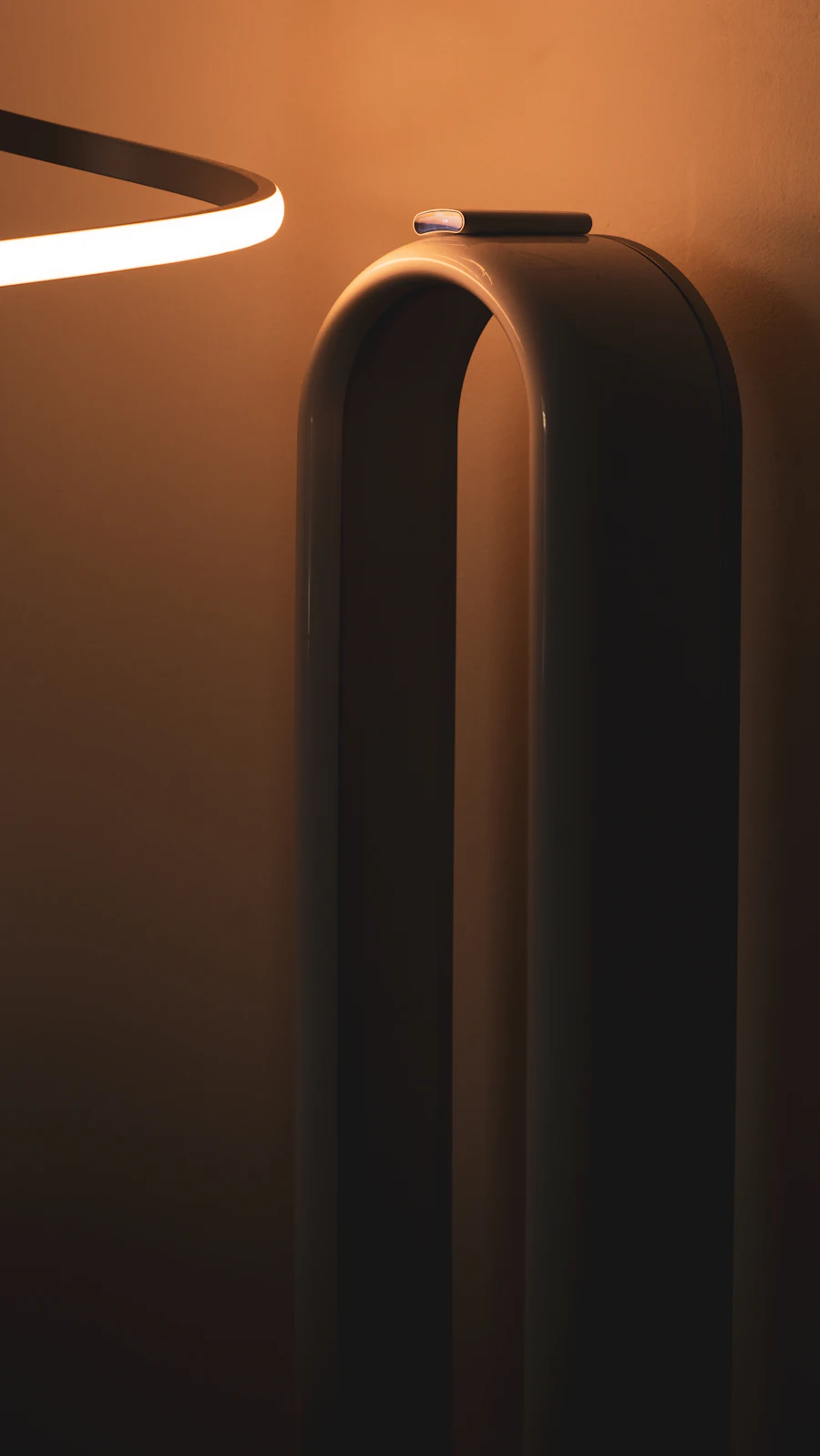 A sleek dark lamp with a glowing curved light bar casts warm illumination across a plain tan wall. The composition emphasizes smooth industrial shapes and soft shadows.