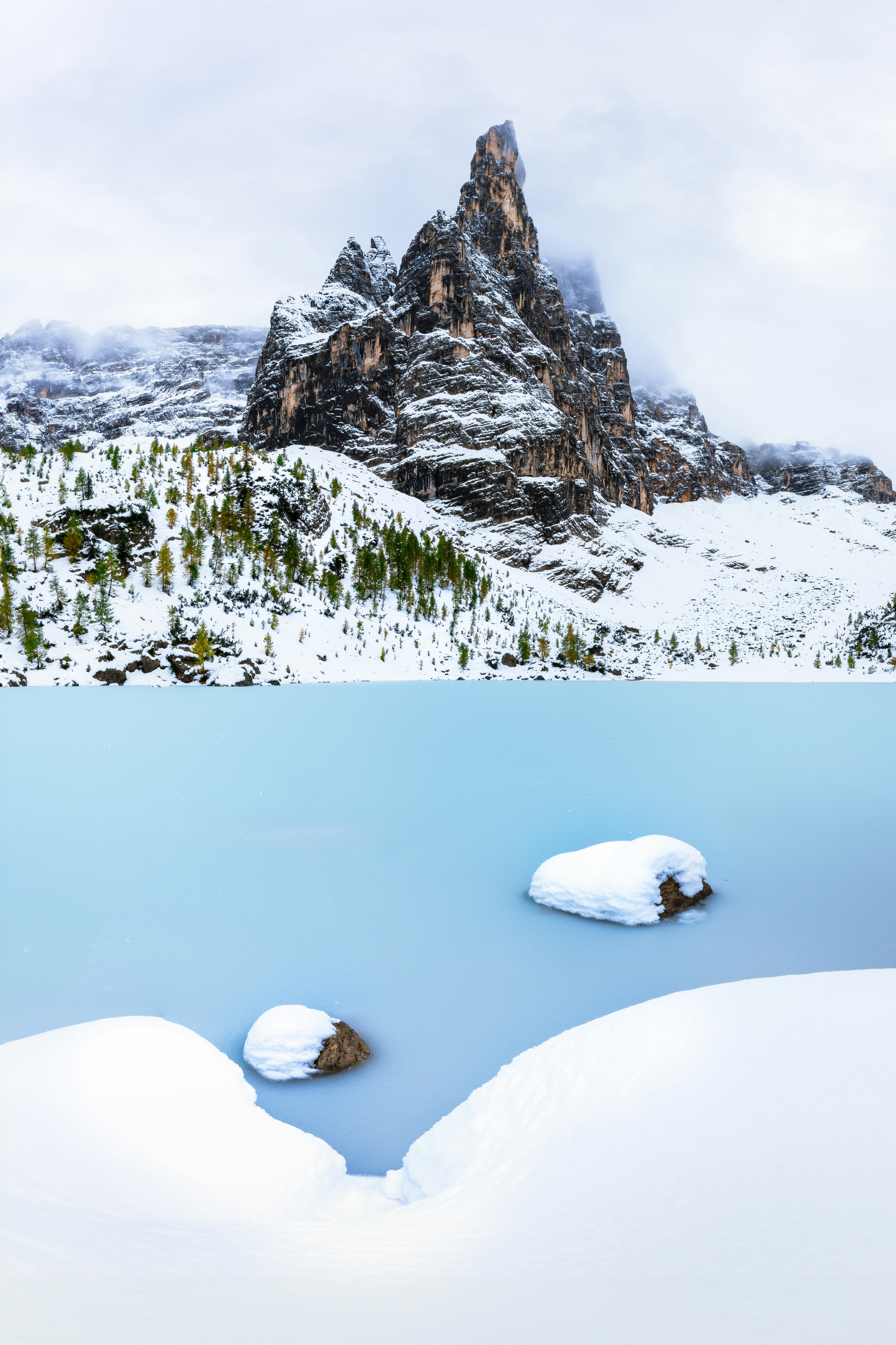 Snow-dusted mountains rise sharply above a serene, ice-blue lake partially covered with snow. Coniferous trees add a touch of green to the wintry scene.