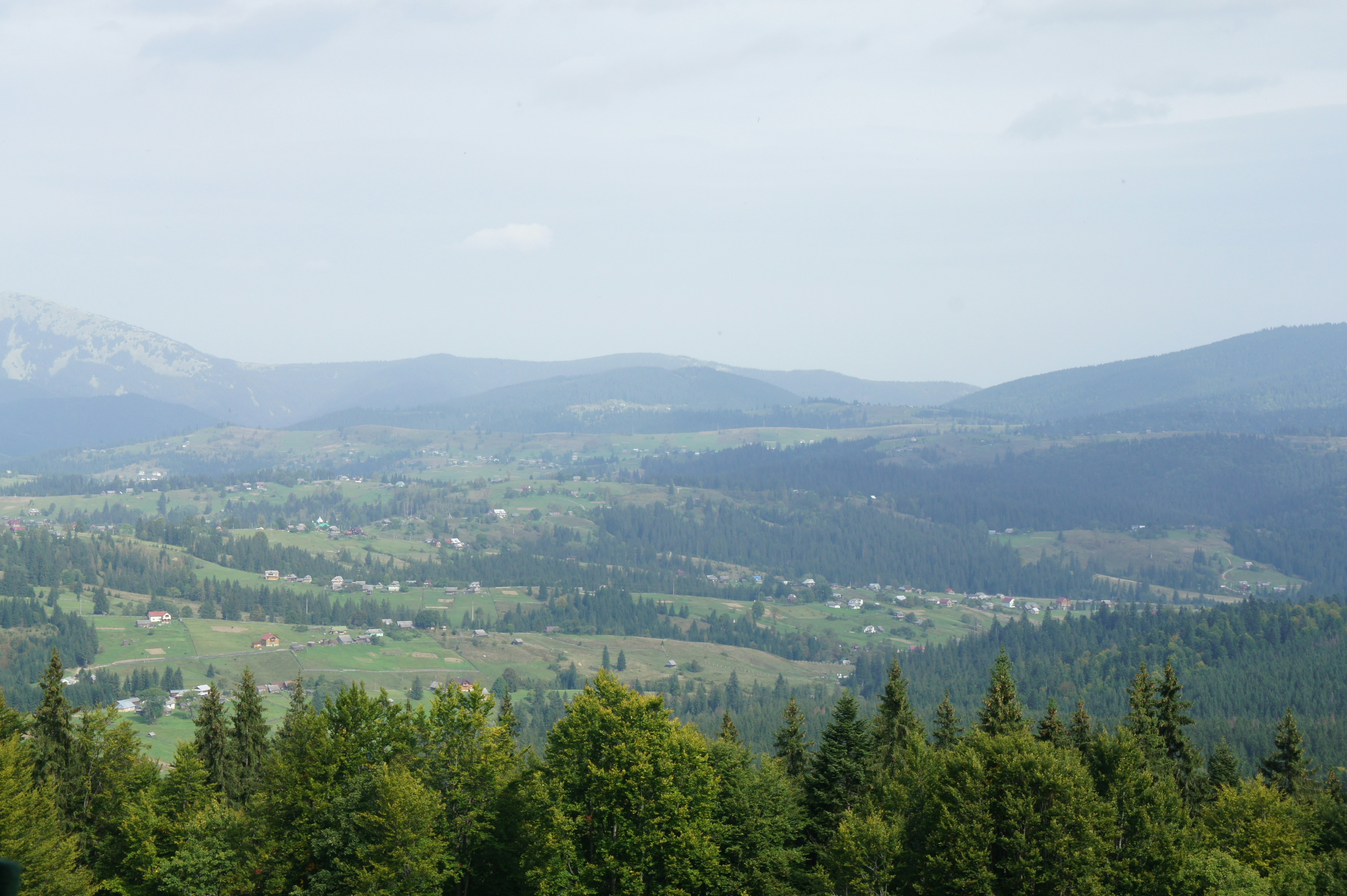 Lush green valleys stretch across the landscape, dotted with quaint houses and framed by distant mountains under a soft, cloudy sky.