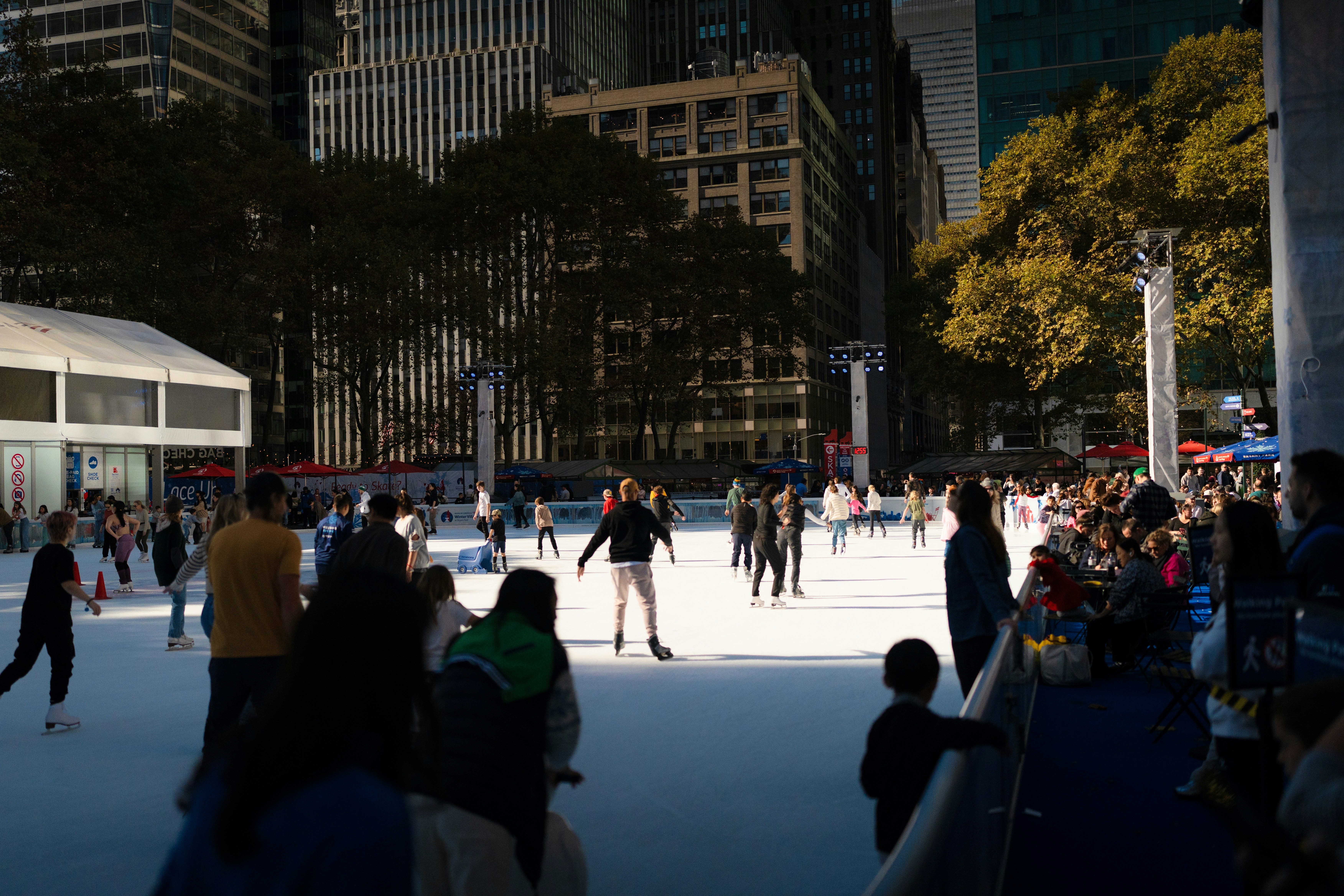Skaters glide across the ice rink in Bryant Park, surrounded by towering skyscrapers and vibrant autumn foliage. A lively gathering of people captures the essence of seasonal festivities.