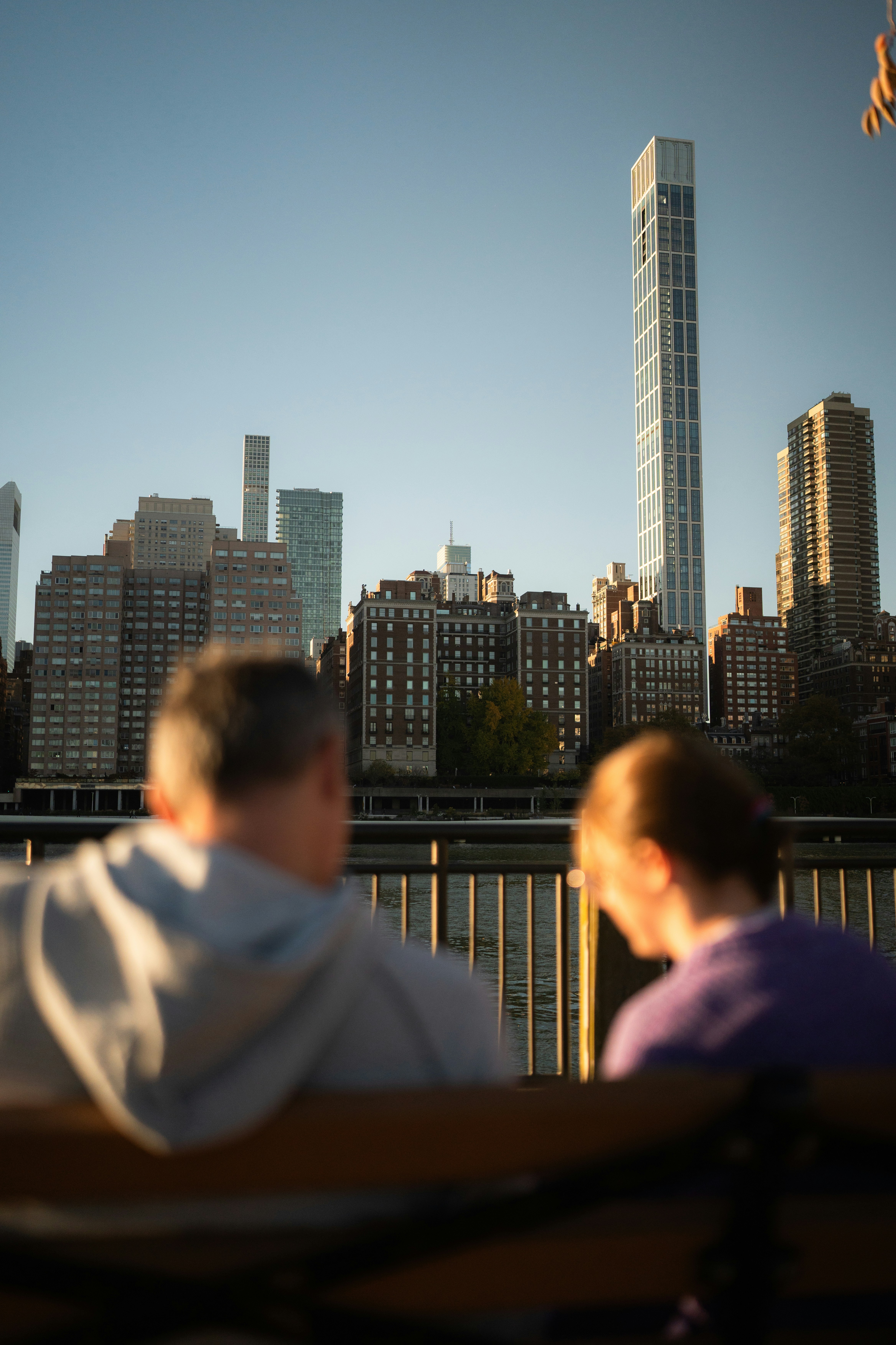 Two individuals sit on a bench in soft focus, framed by a city skyline under a clear blue sky, with warm sunlight highlighting architectural details.
