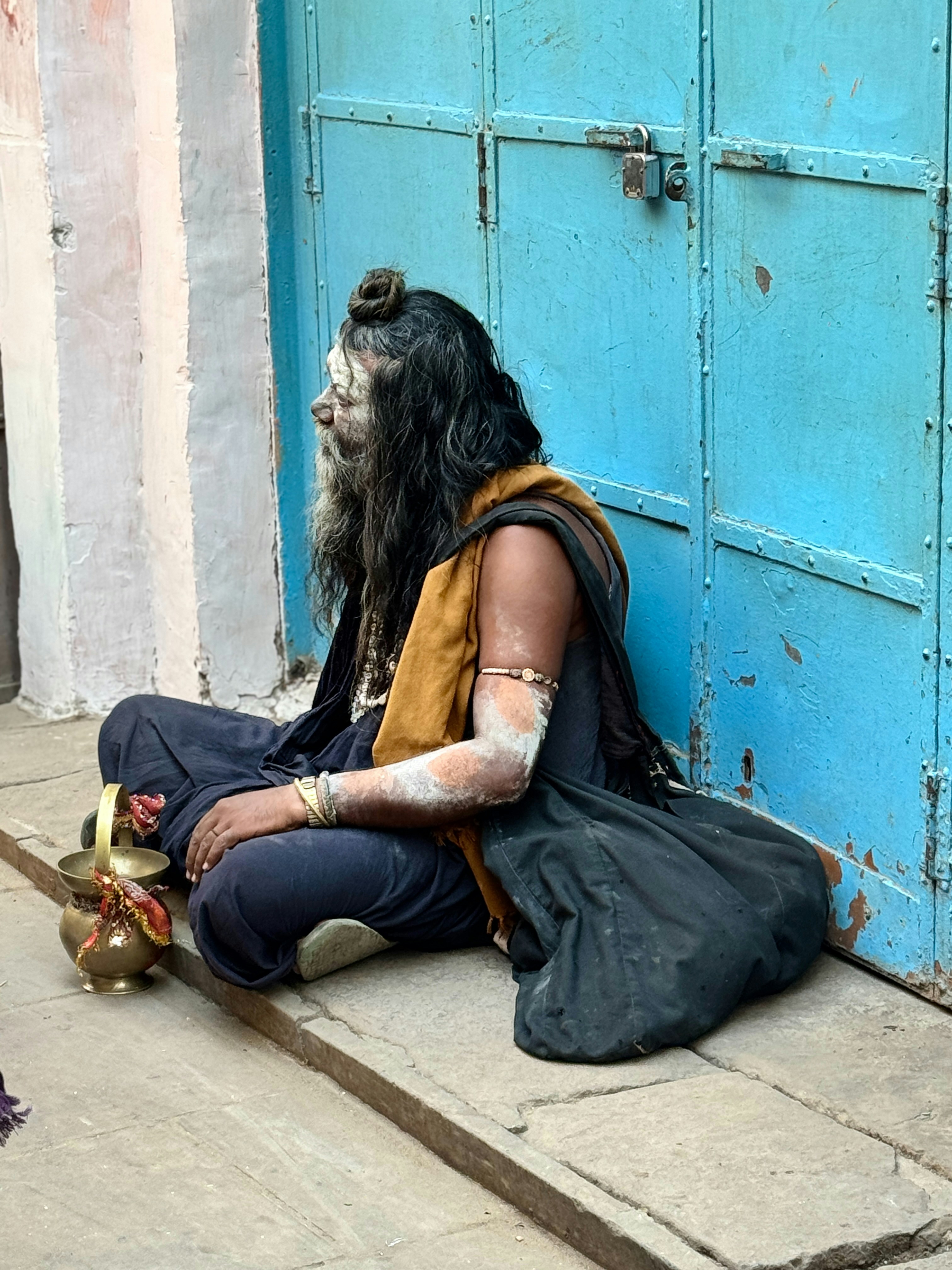 Ash-smeared sadhu in front of turquoise doors with brass offering pot