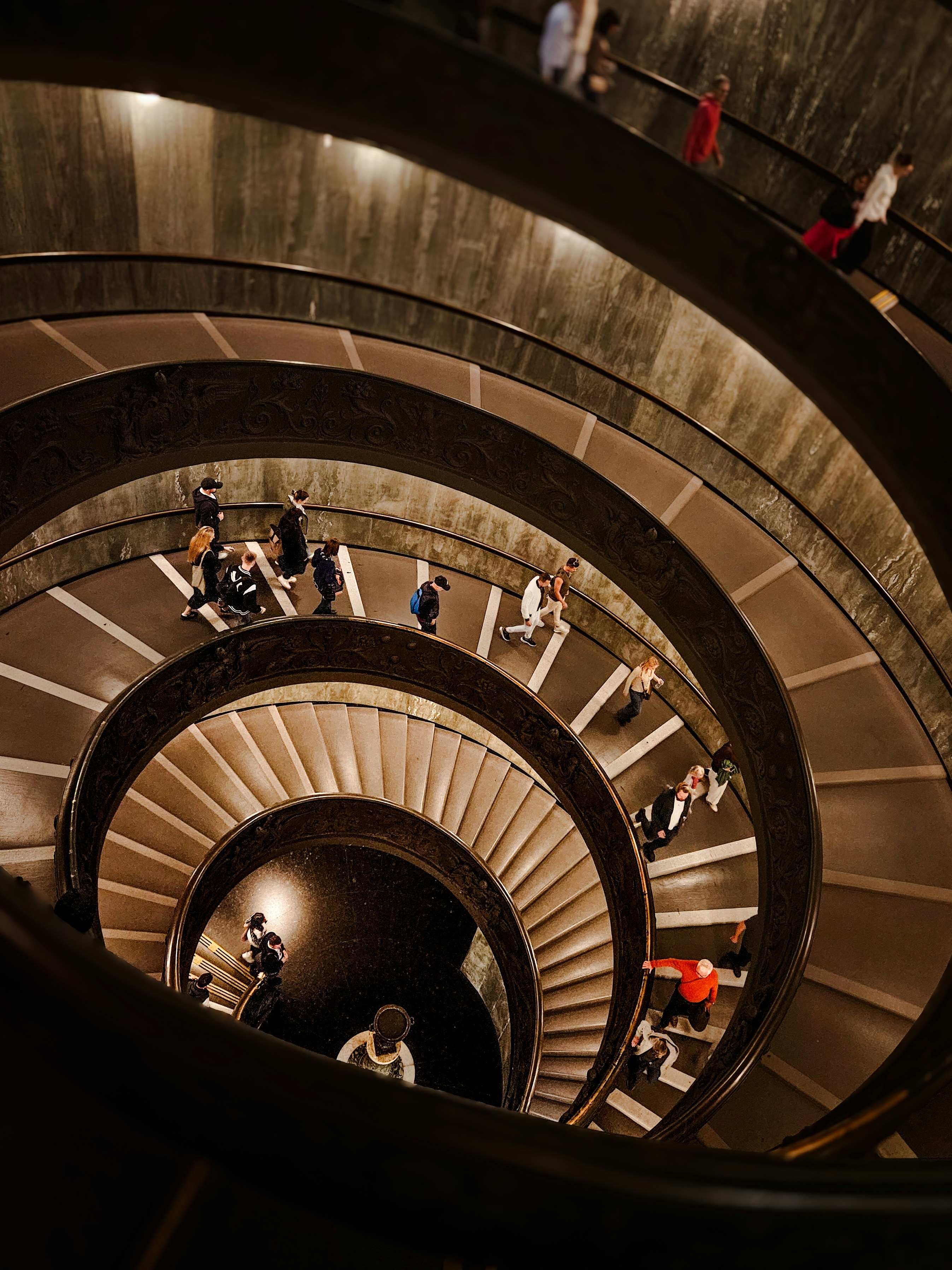 Overhead view of a grand spiral staircase, with visitors tracing the ornate railings as concentric circles descend toward a dim central void.