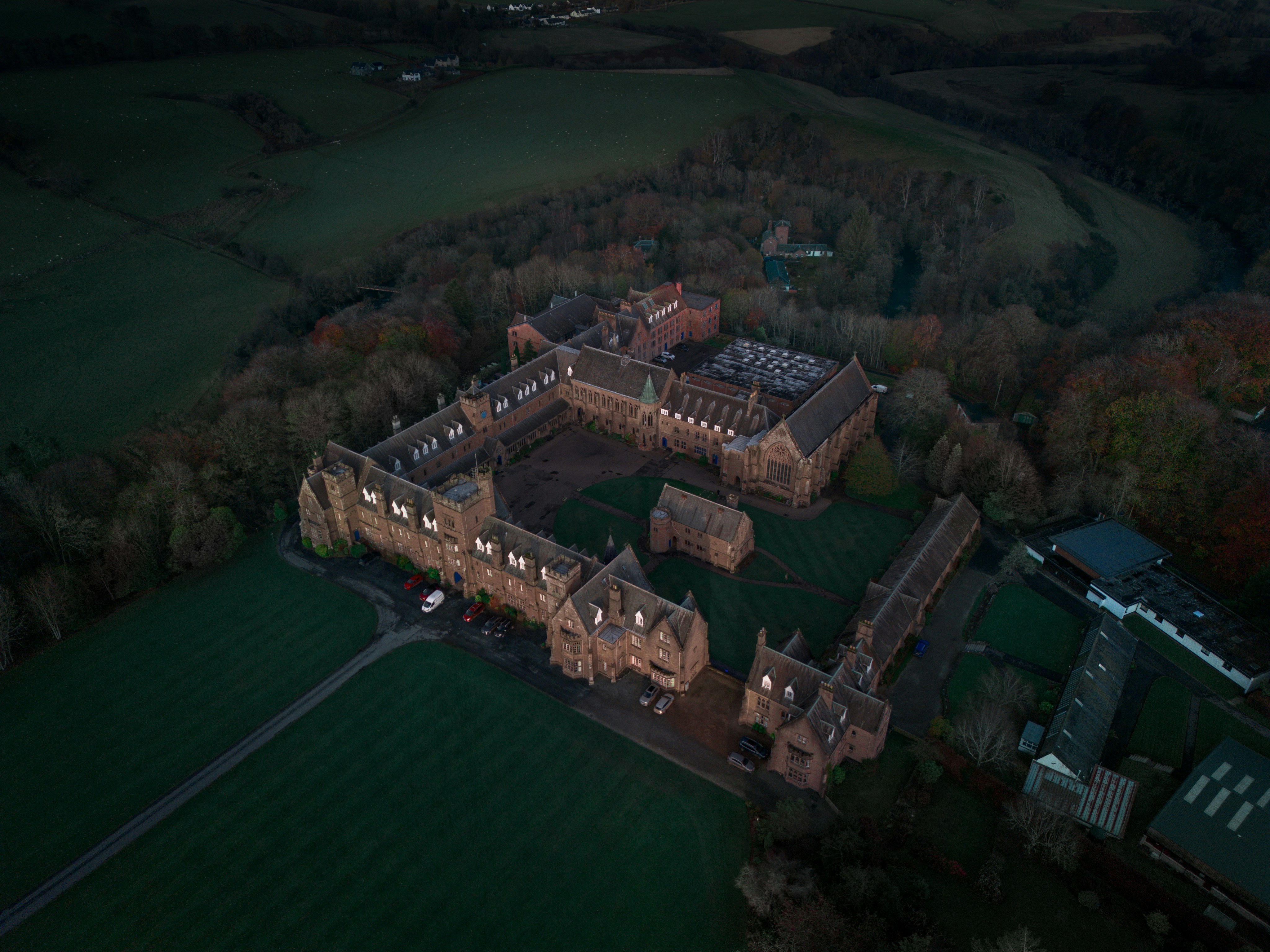 Drone-captured aerial view of a historic castle complex with a central courtyard, framed by autumnal woods and open fields. The symmetrical layout and crenellated walls stand out under subdued evening light.