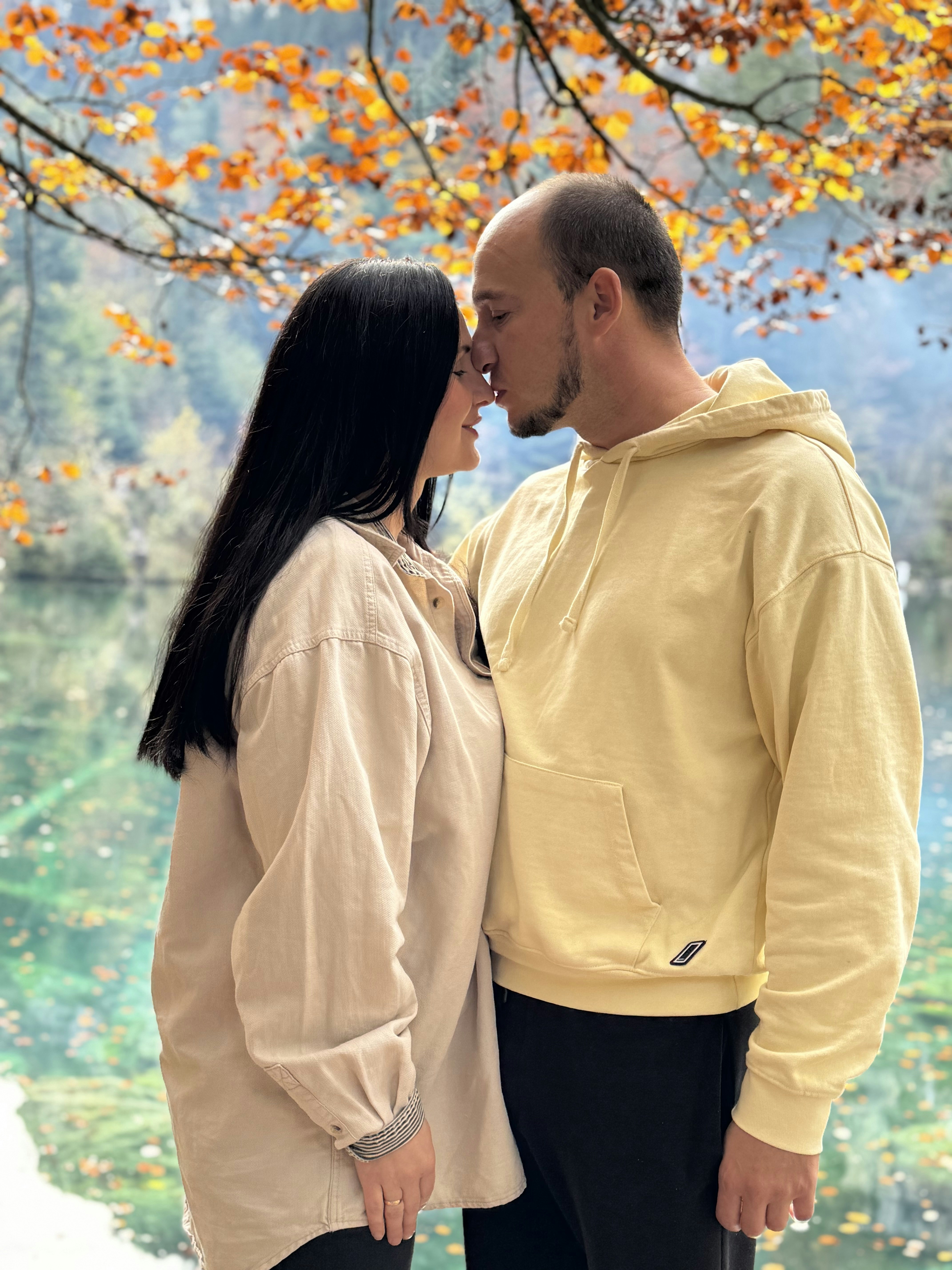 Couple sharing an intimate moment with foreheads touching, surrounded by vibrant autumn leaves and a serene lake backdrop.