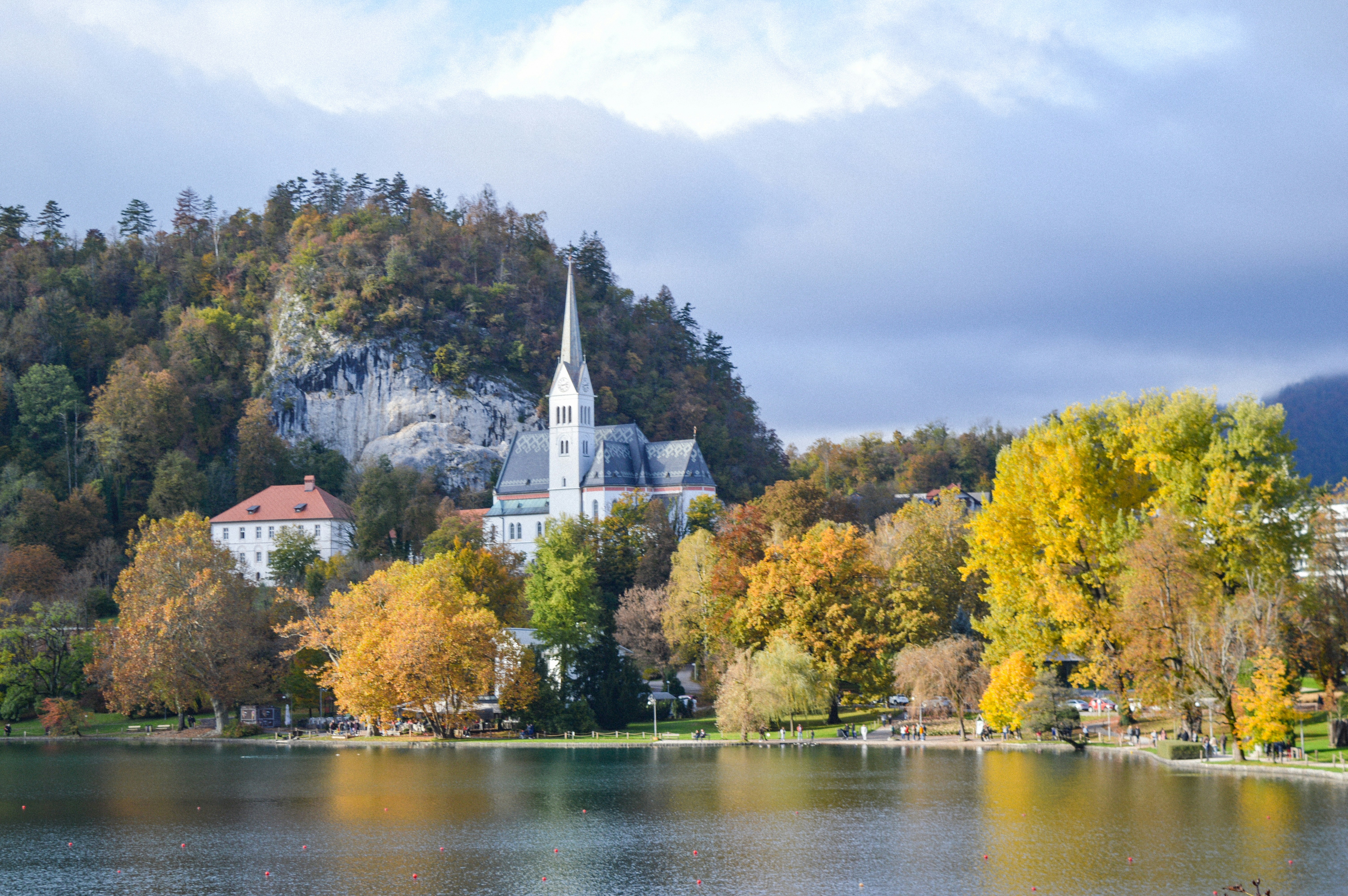 Chapel with a tall spire nestled among colorful autumn trees by a tranquil lake.