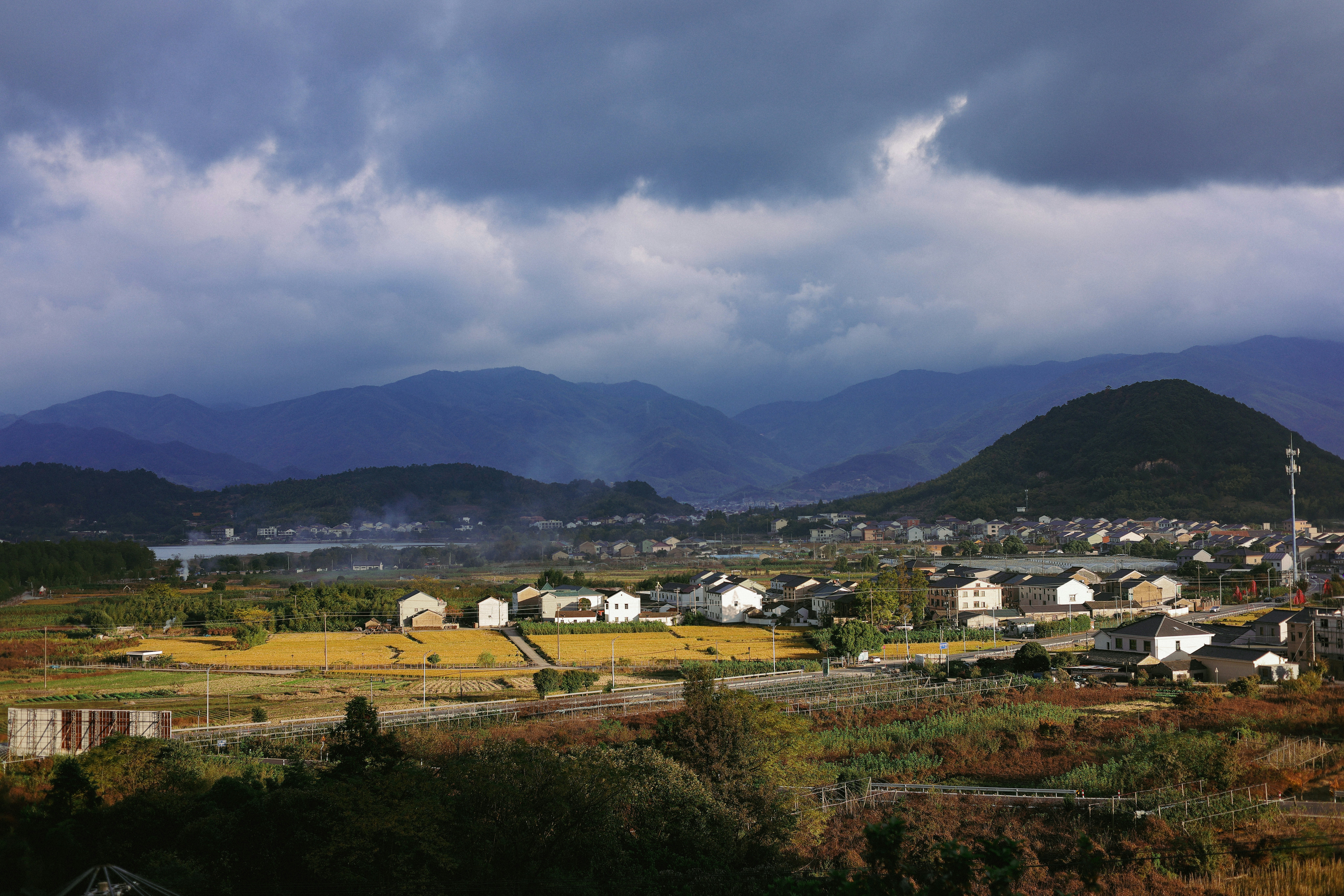 A serene landscape showcasing agricultural fields and quaint houses nestled against a backdrop of rolling mountains under a dramatic sky.
