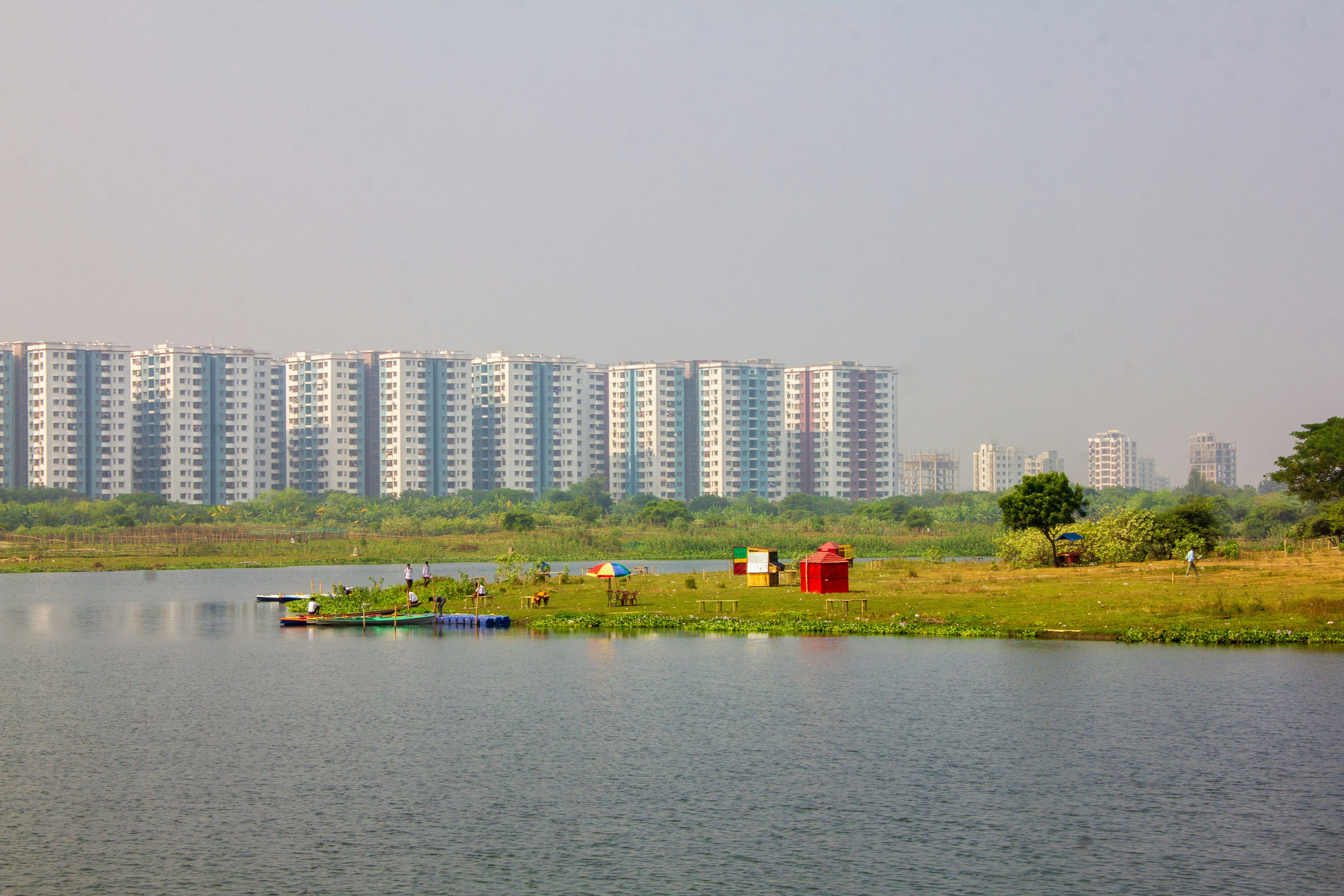 Expansive view of a serene lake bordered by modern high-rise buildings, with a small colorful structure and greenery in the foreground.