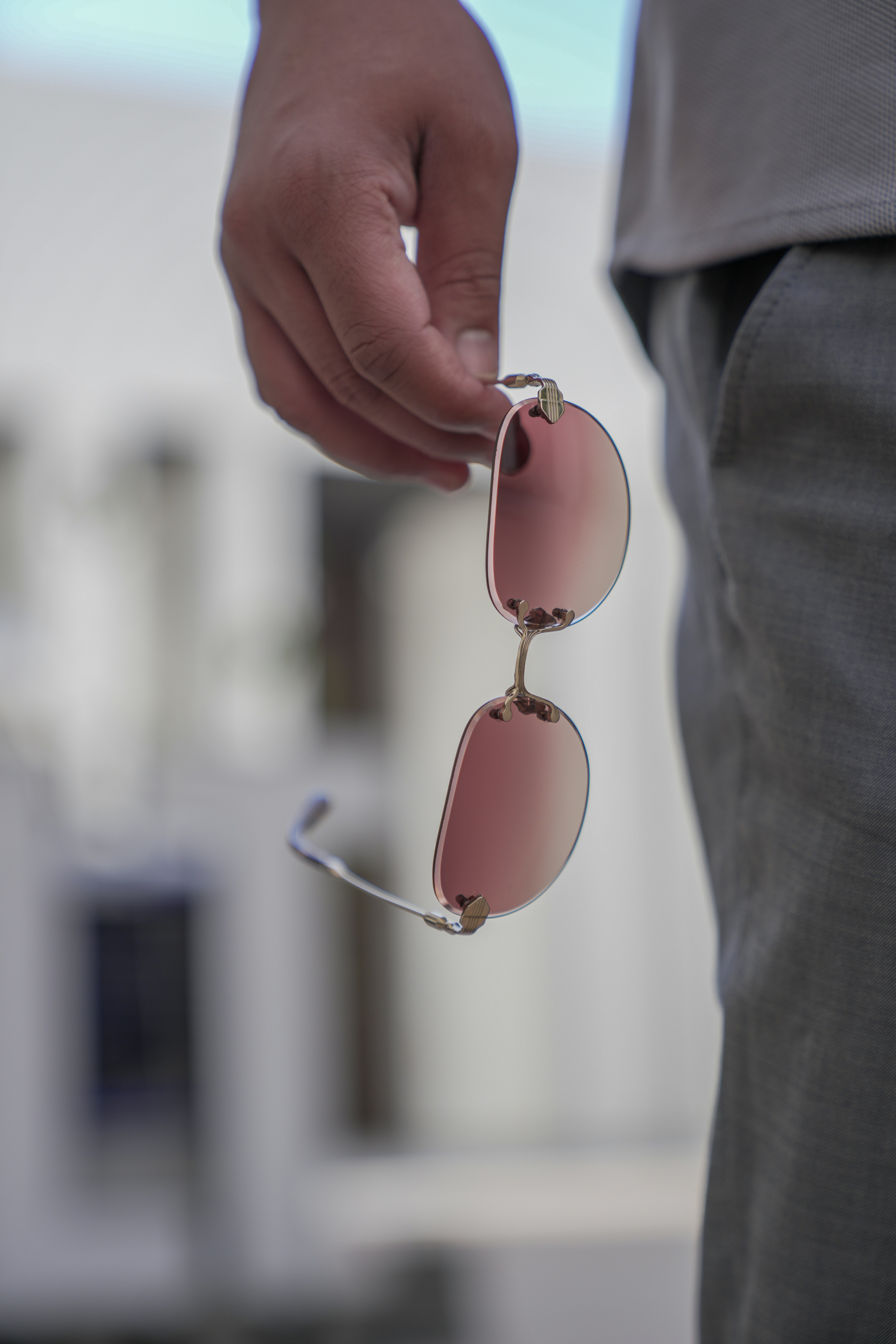 Pink-tinted sunglasses dangle from a gloved grip, against a blurred urban backdrop