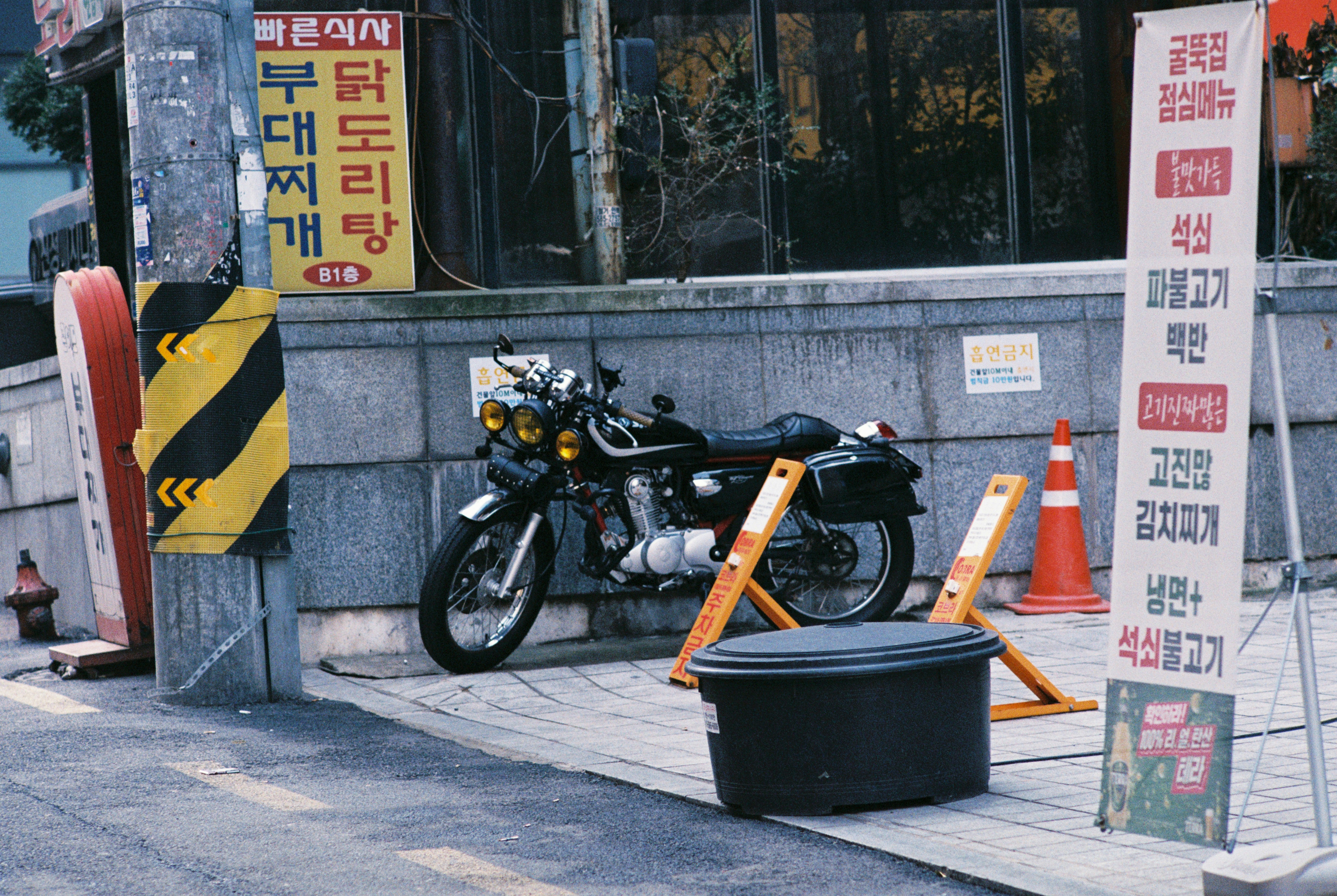 Motorcycle parked beside a concrete wall with orange barriers and traffic cones. Korean storefront signs loom in the background.
