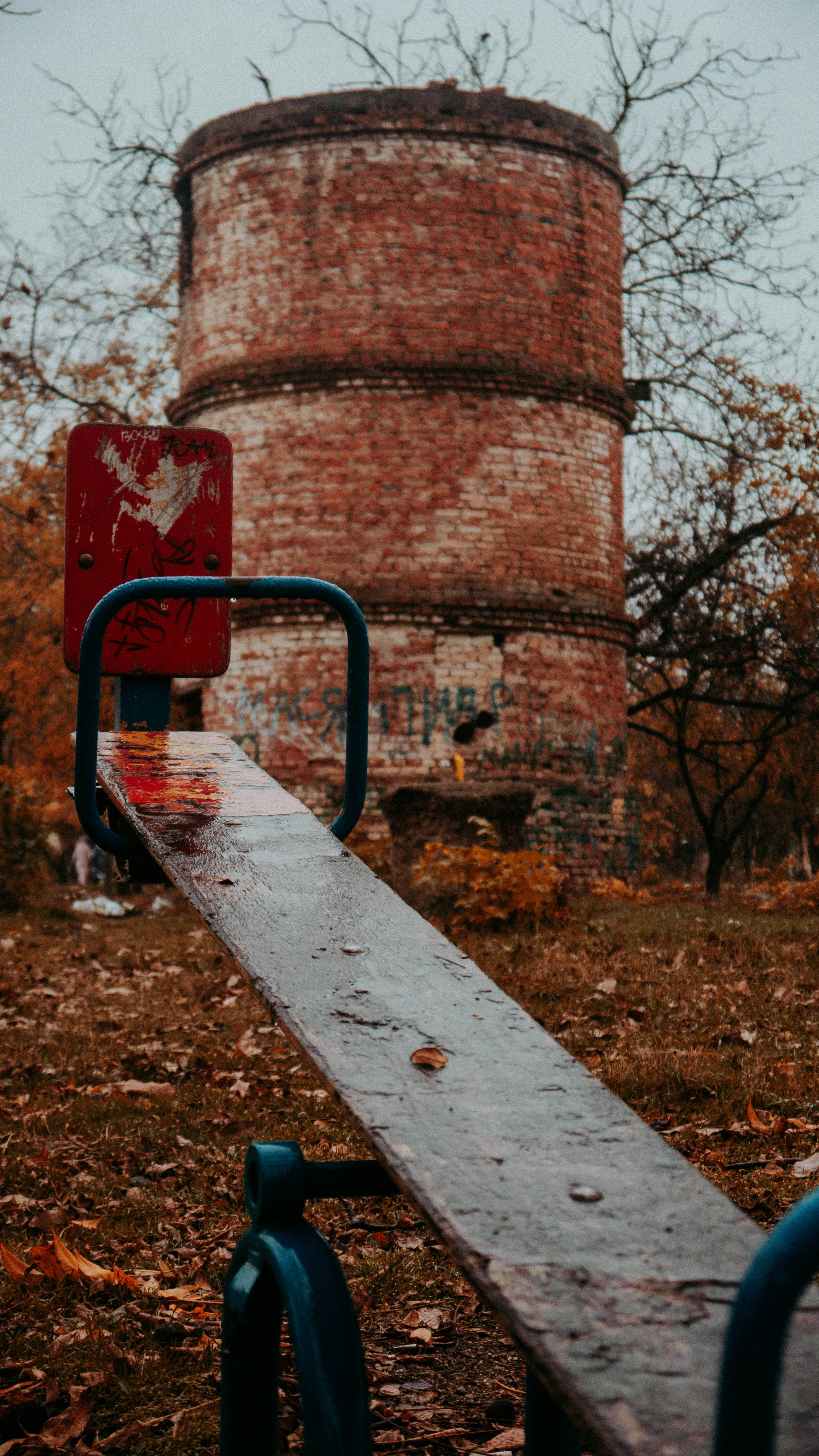 Weathered playground seesaw beam extends toward a tall circular brick tower in an autumn park.