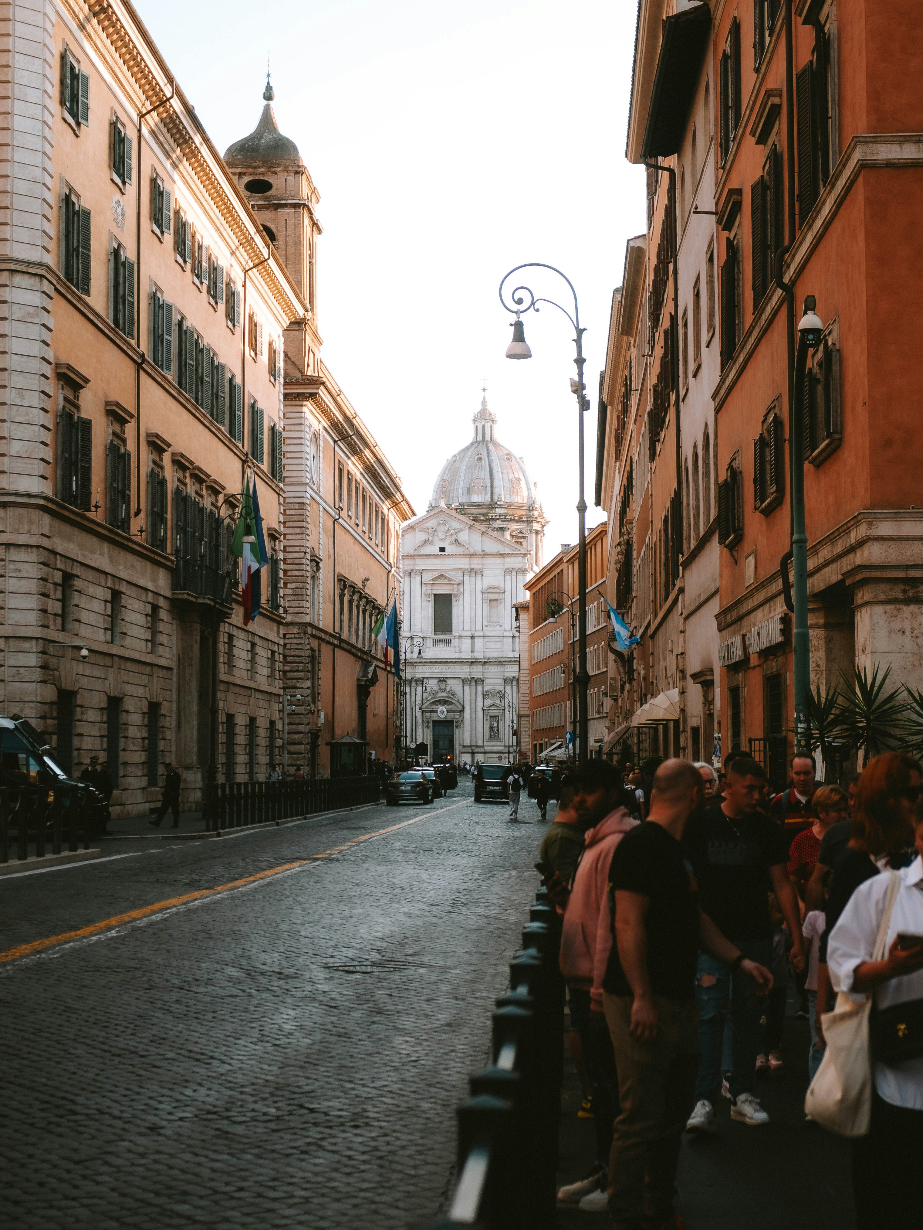 The image appears to be a street scene in Rome, Italy. The image features a street lined with buildings, with people walking along the sidewalk.