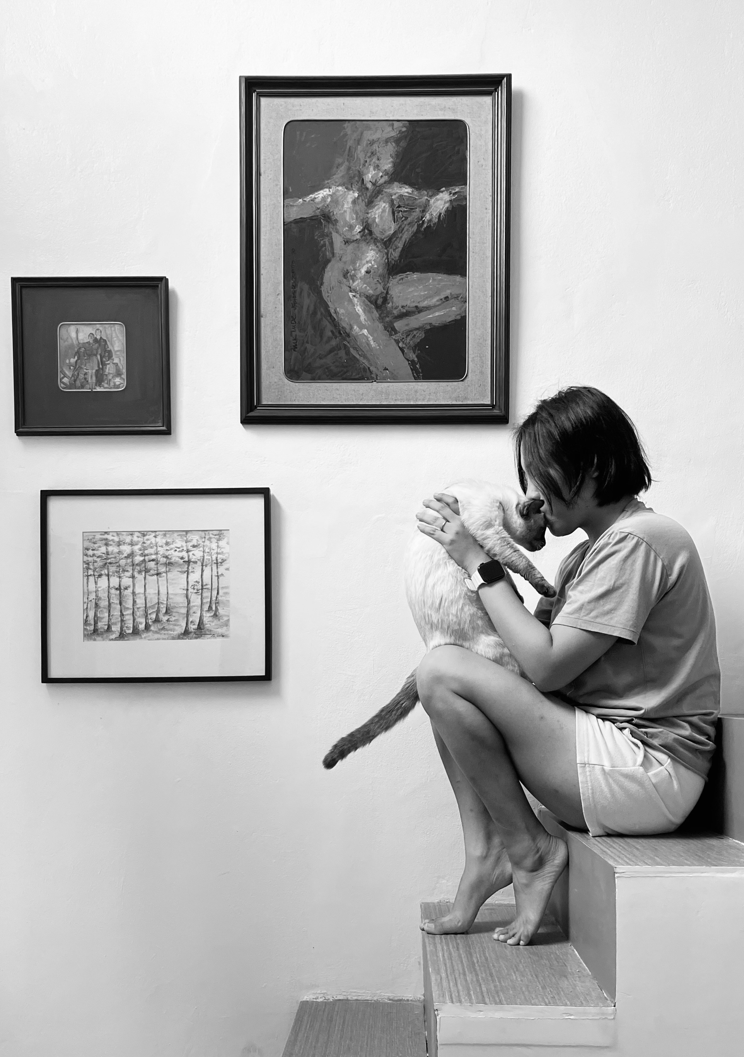 A woman sits on stairs, cradling a cat in her arms, surrounded by framed artworks on the wall. The scene captures a quiet moment of connection.