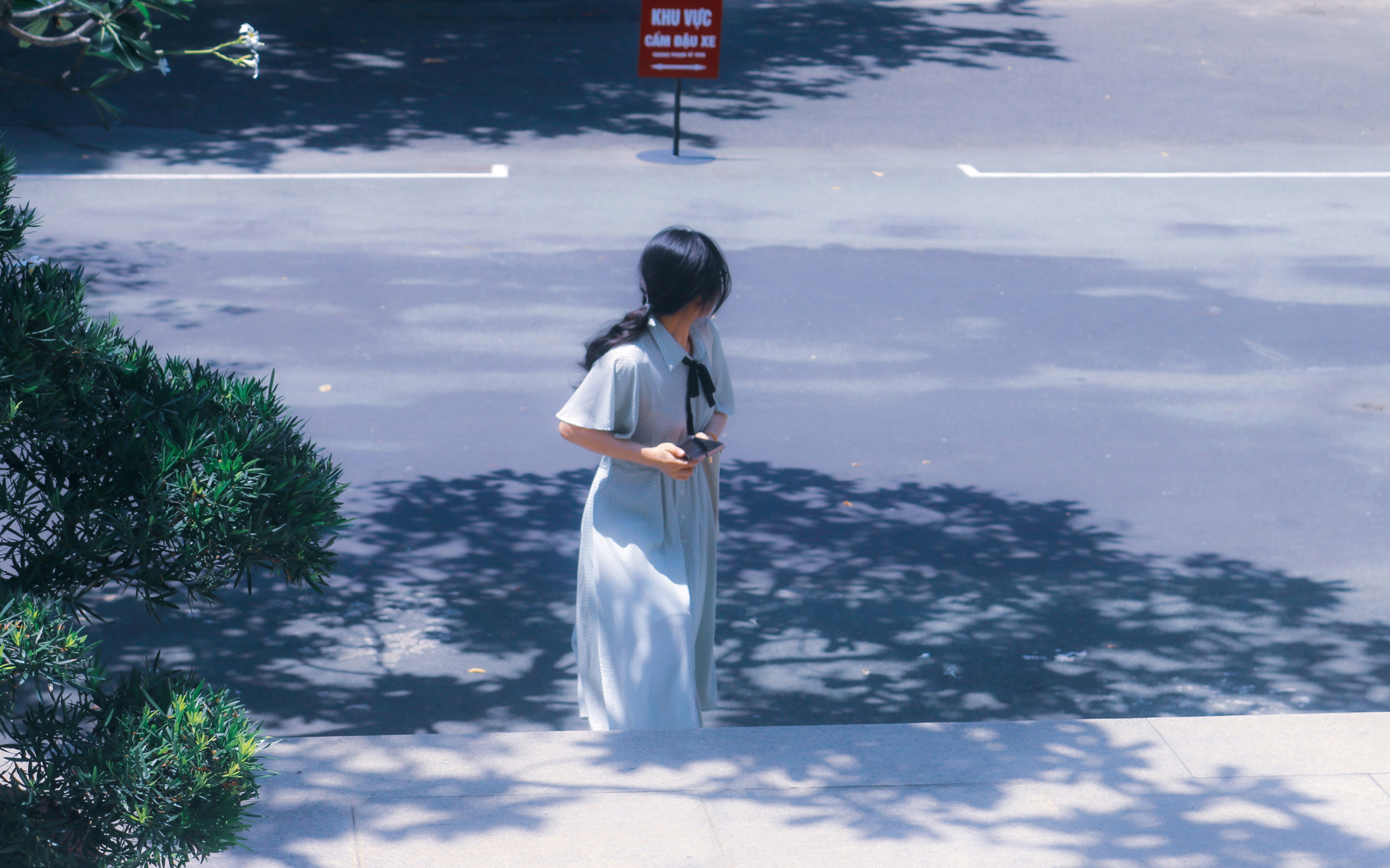 A woman in a flowing gray dress stands on a sunlit street, surrounded by dappled shadows from nearby trees. The scene captures a moment of quiet reflection amidst urban life.