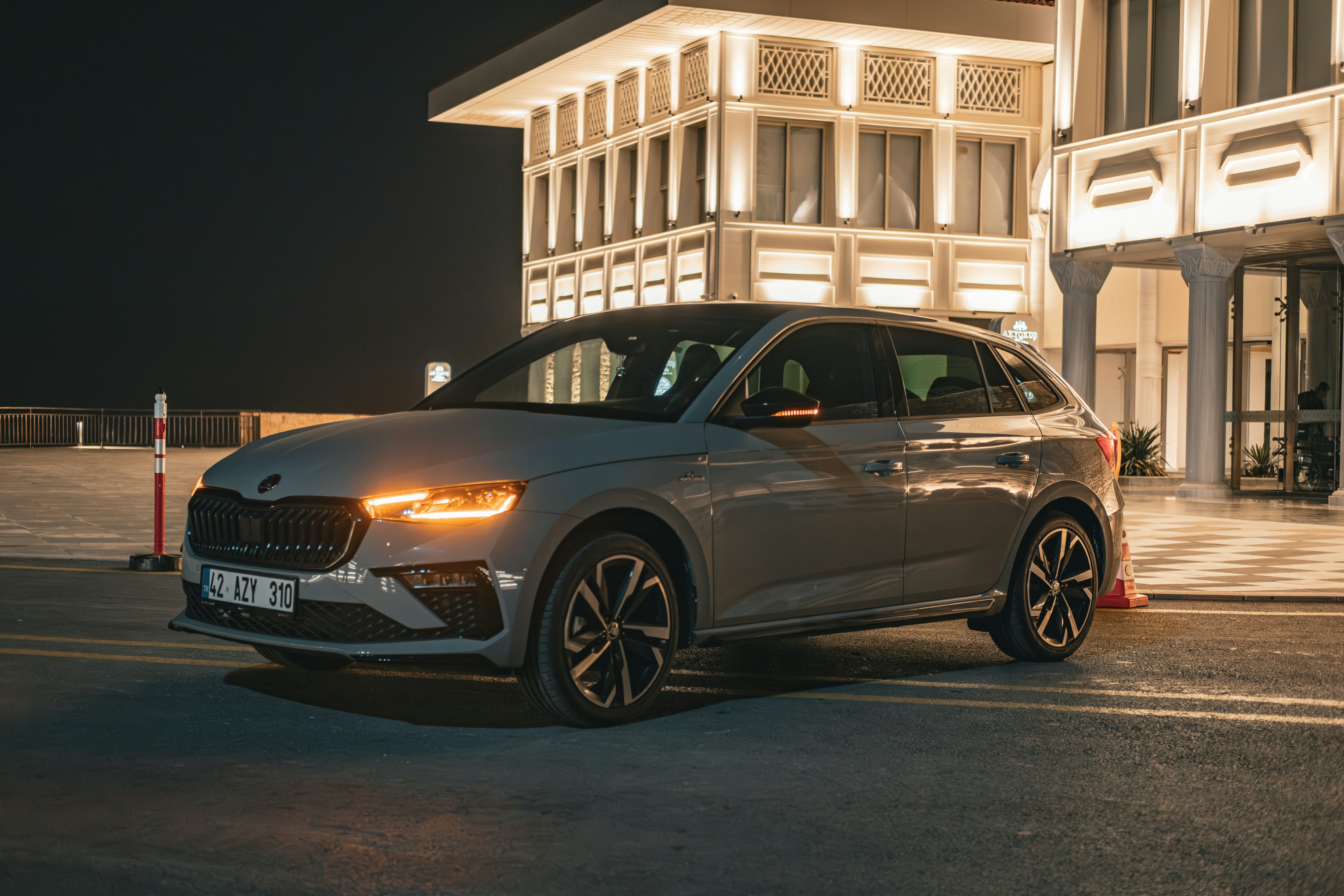A silver-gray car with illuminated headlights is parked in front of a beautifully lit modern building at night