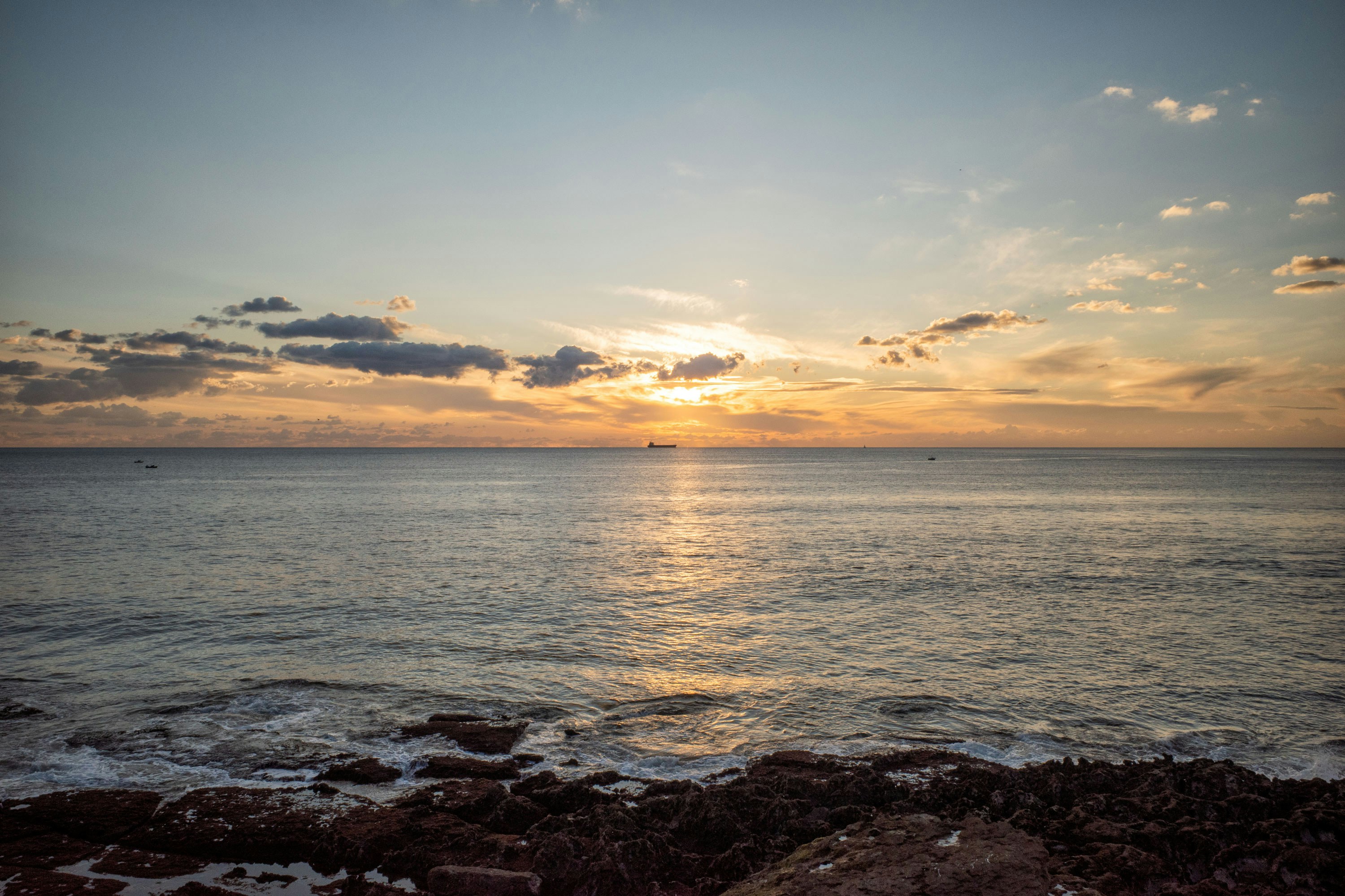 Cargo ship on the horizon at sunset viewed from a rocky shoreline in Portugal.