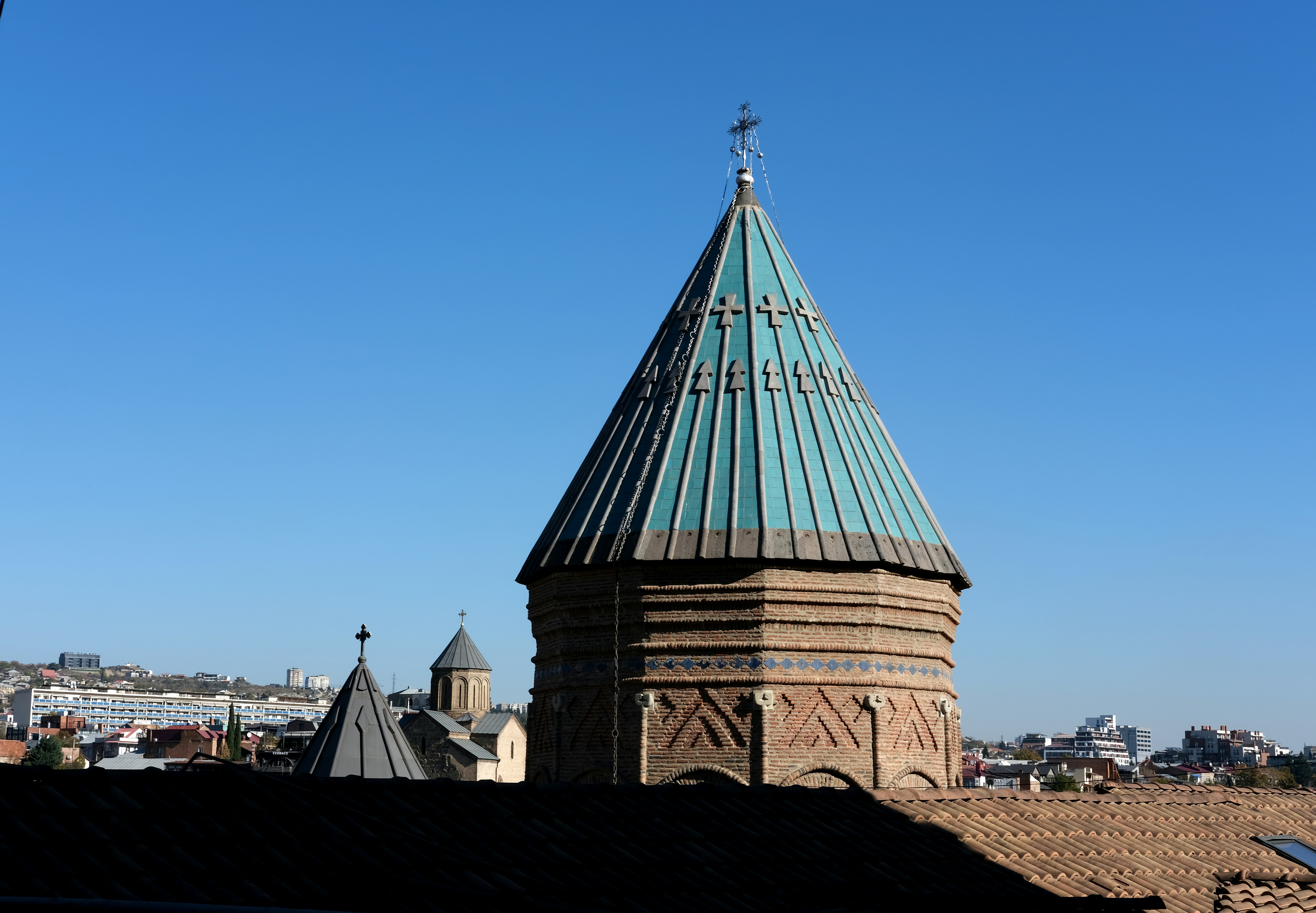 A historical church dome with intricate geometric patterns, captured against the backdrop of a bright blue sky and a distant urban landscape. The conical shape of the roof, adorned with turquoise details and crosses, adds to the architectural charm of this cultural heritage site. The juxtaposition of modern city elements in the background highlights the harmony between old and new.