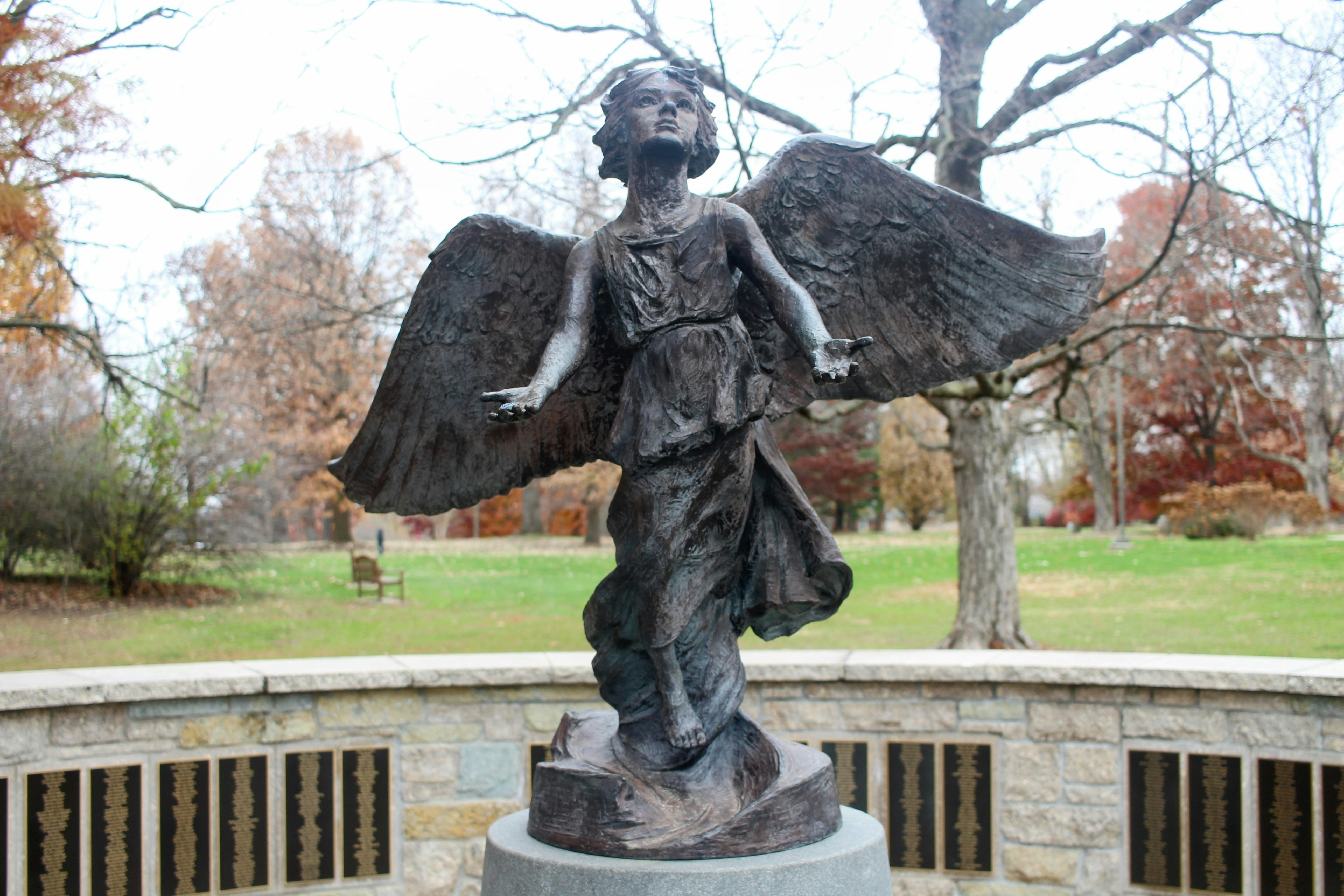 Bronze statue of a woman with large wings set in a serene park setting during autumn.