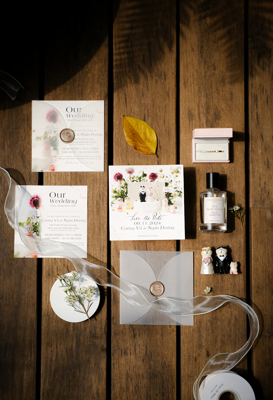 Elegant wedding invitation suite arranged on a wooden plank surface with ribbon, wax seals, flowers, perfume, and small figurines. Warm sunlight casts soft shadows across the stationery.