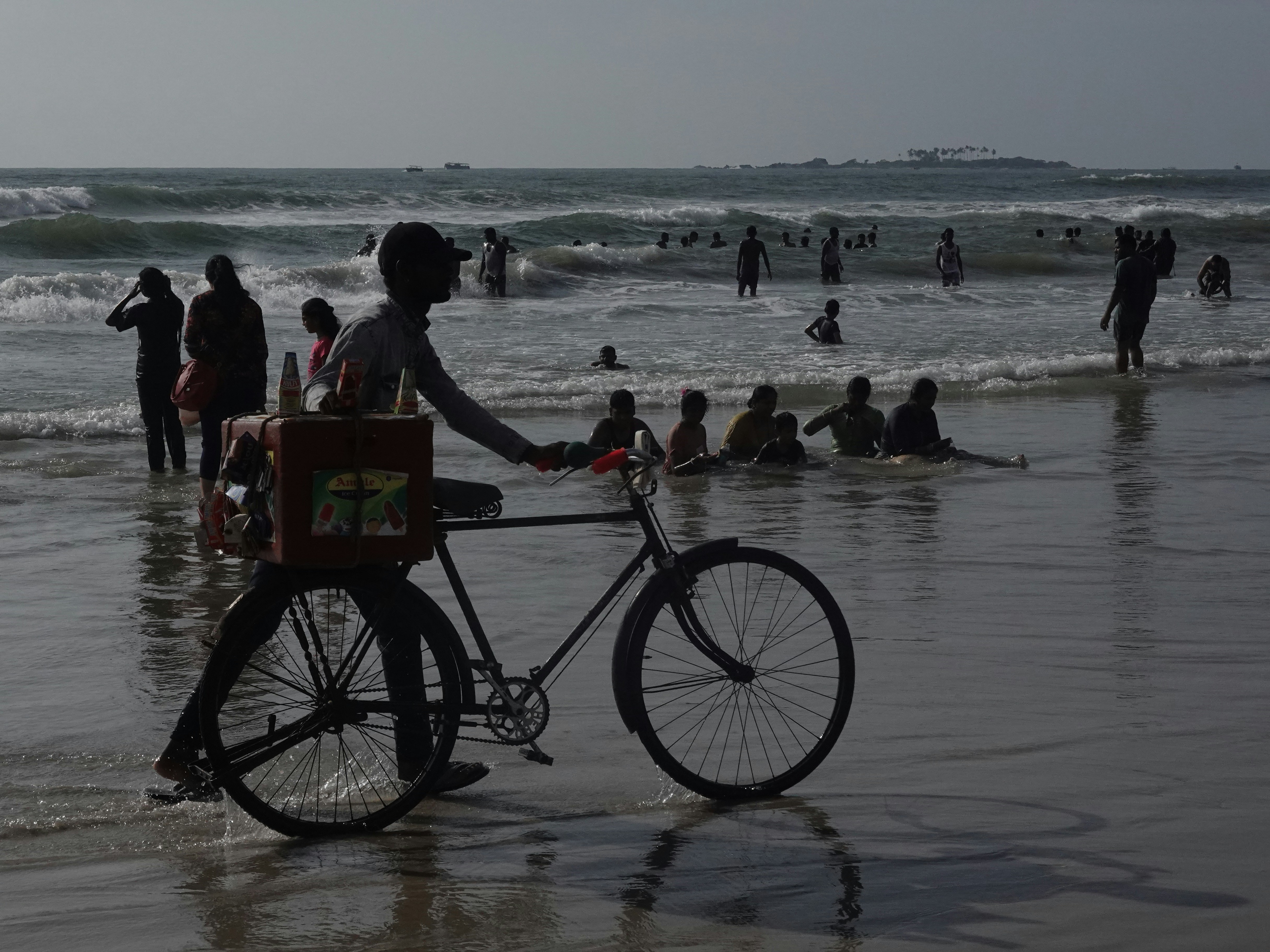 Silhouetted figures on a beach with a cyclist pushing a bicycle along the shoreline as others swim in the ocean.