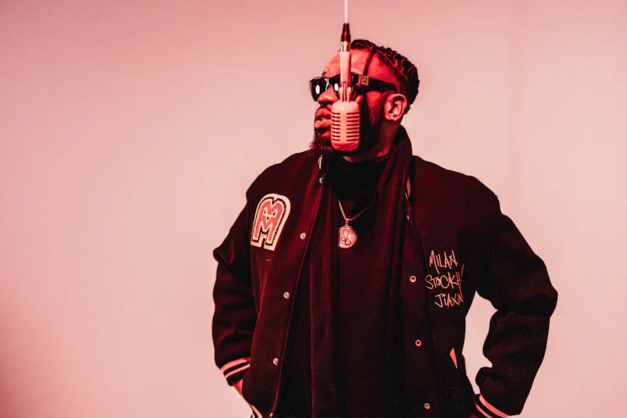 A man in sunglasses and a dark varsity jacket poses under a vintage hanging microphone against a pink-lit backdrop.