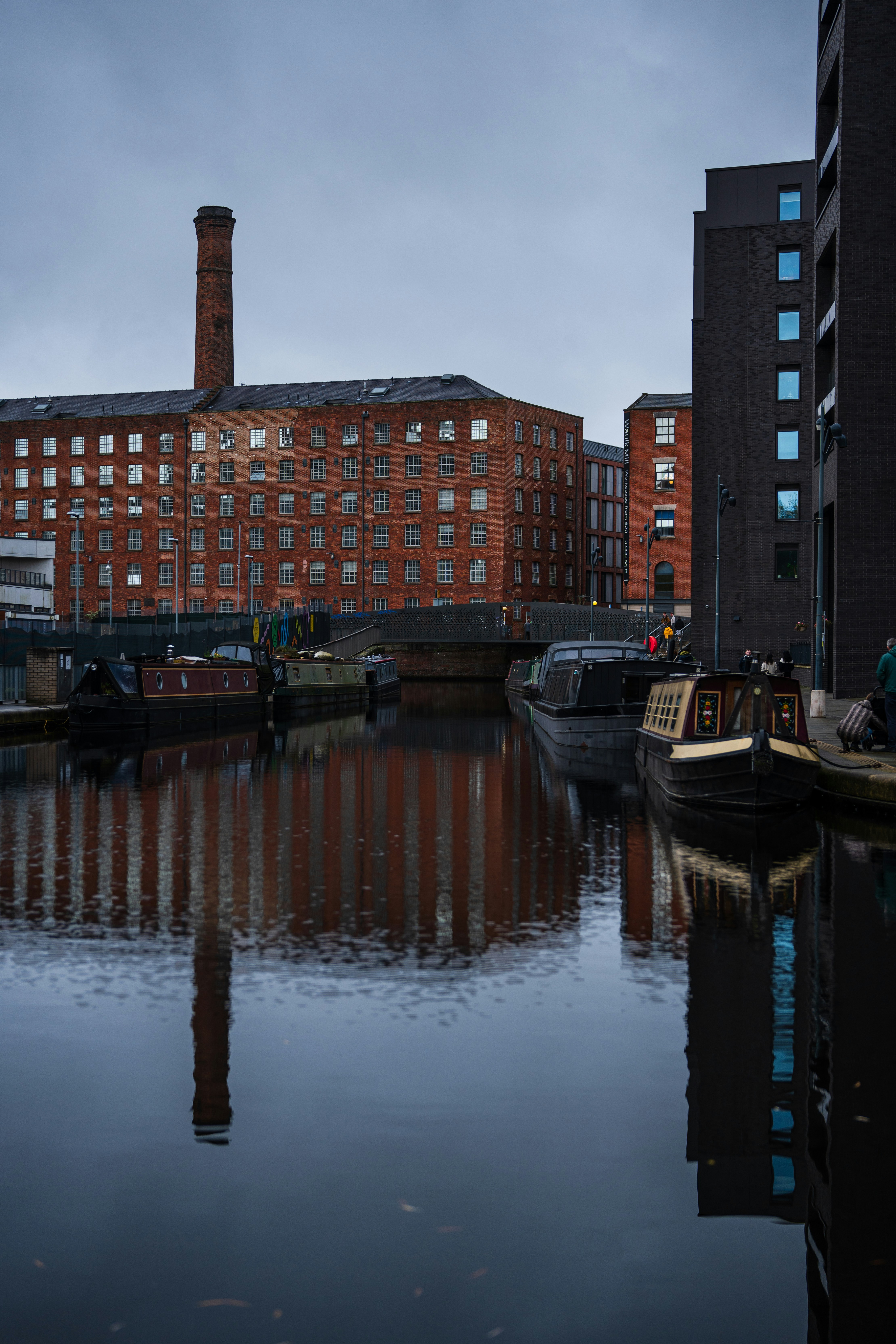 Historic red brick warehouse reflected in calm water, with moored boats and modern buildings in the foreground.