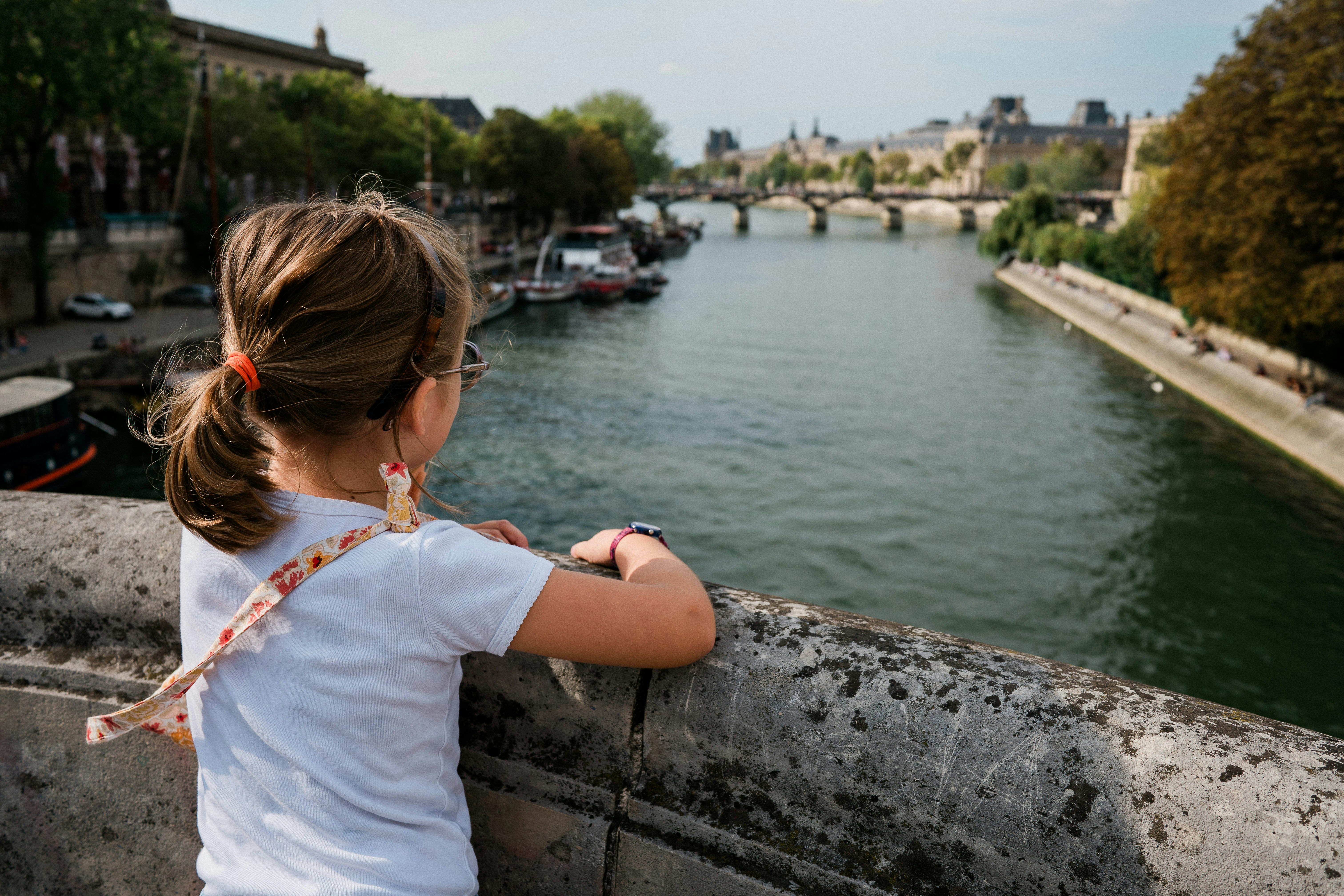 A young girl rests her arms on a stone railing, looking toward a sunlit river with boats and arched bridges. The cityscape photograph captures a quiet moment from the bridge.