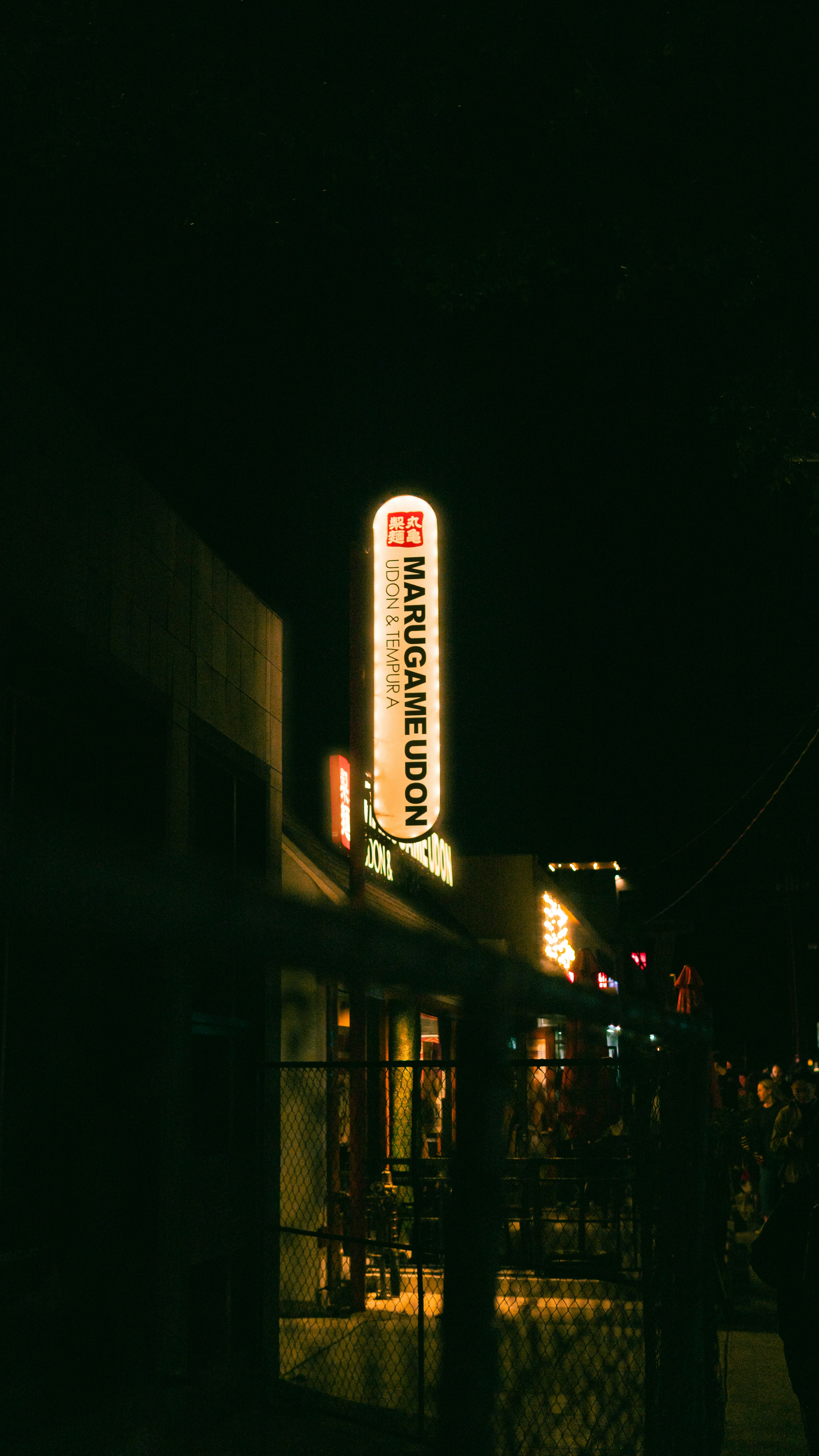 Vertical Neon: Marugame Udon Rises Over a Fence-Lined Night Street