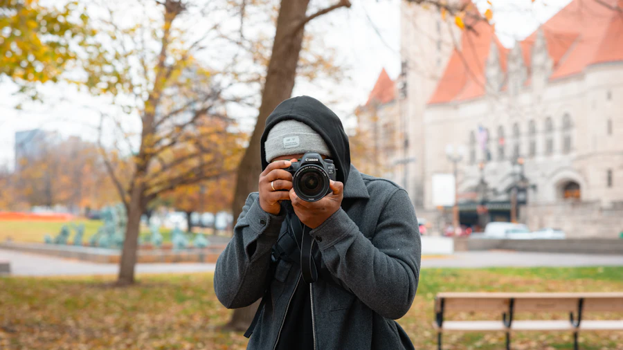 Photographer in a gray coat aiming a camera toward the viewer amid autumn park trees and a historic building backdrop.