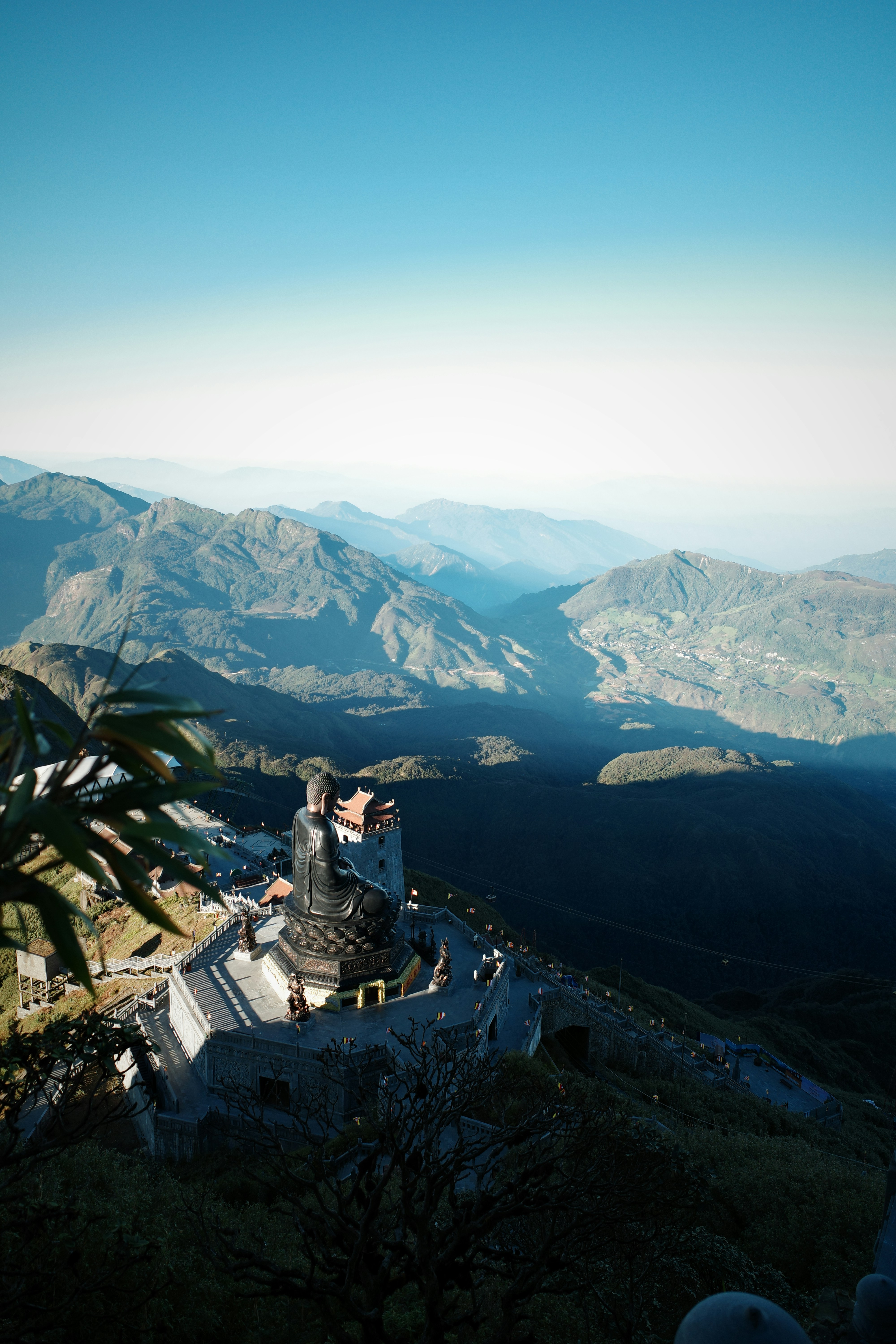 Majestic statue of a seated Buddha overlooking a vast mountain range, with visitors exploring the surrounding area. The scene captures a serene moment in a breathtaking landscape.