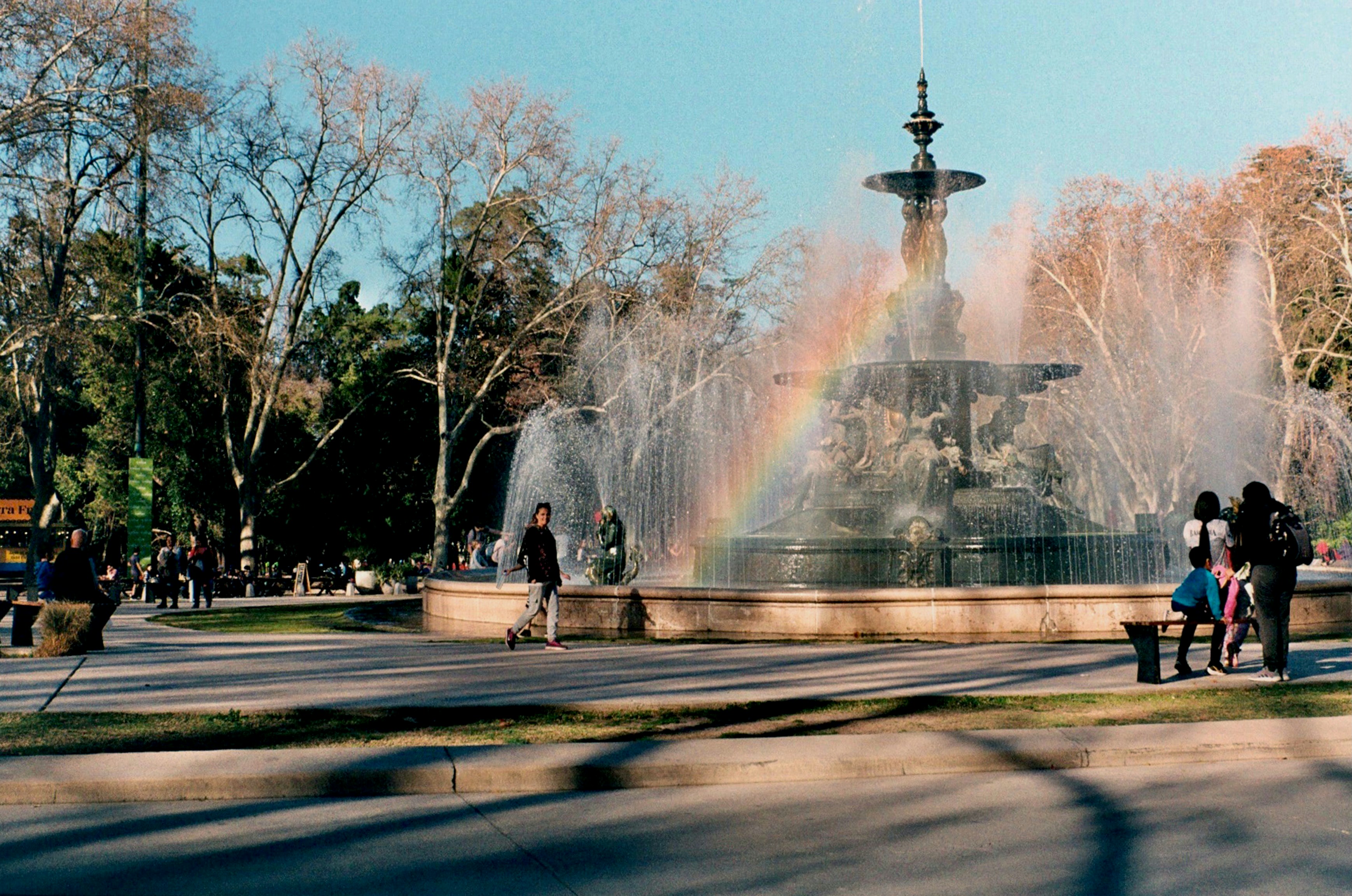 A group of people standing around a fountain with a rainbow in the background