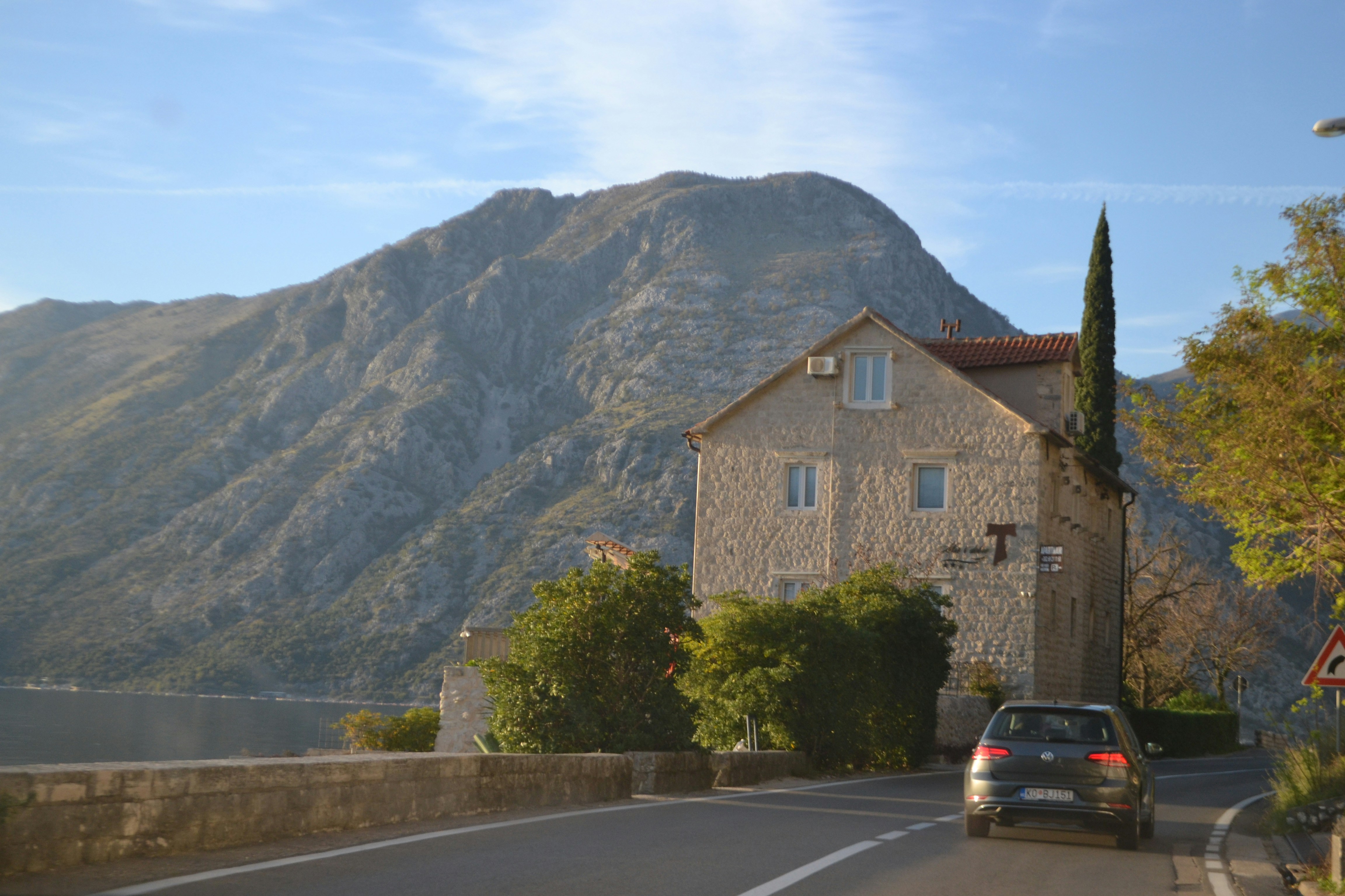 Historic stone house by a winding road with a majestic mountain backdrop under a clear blue sky.