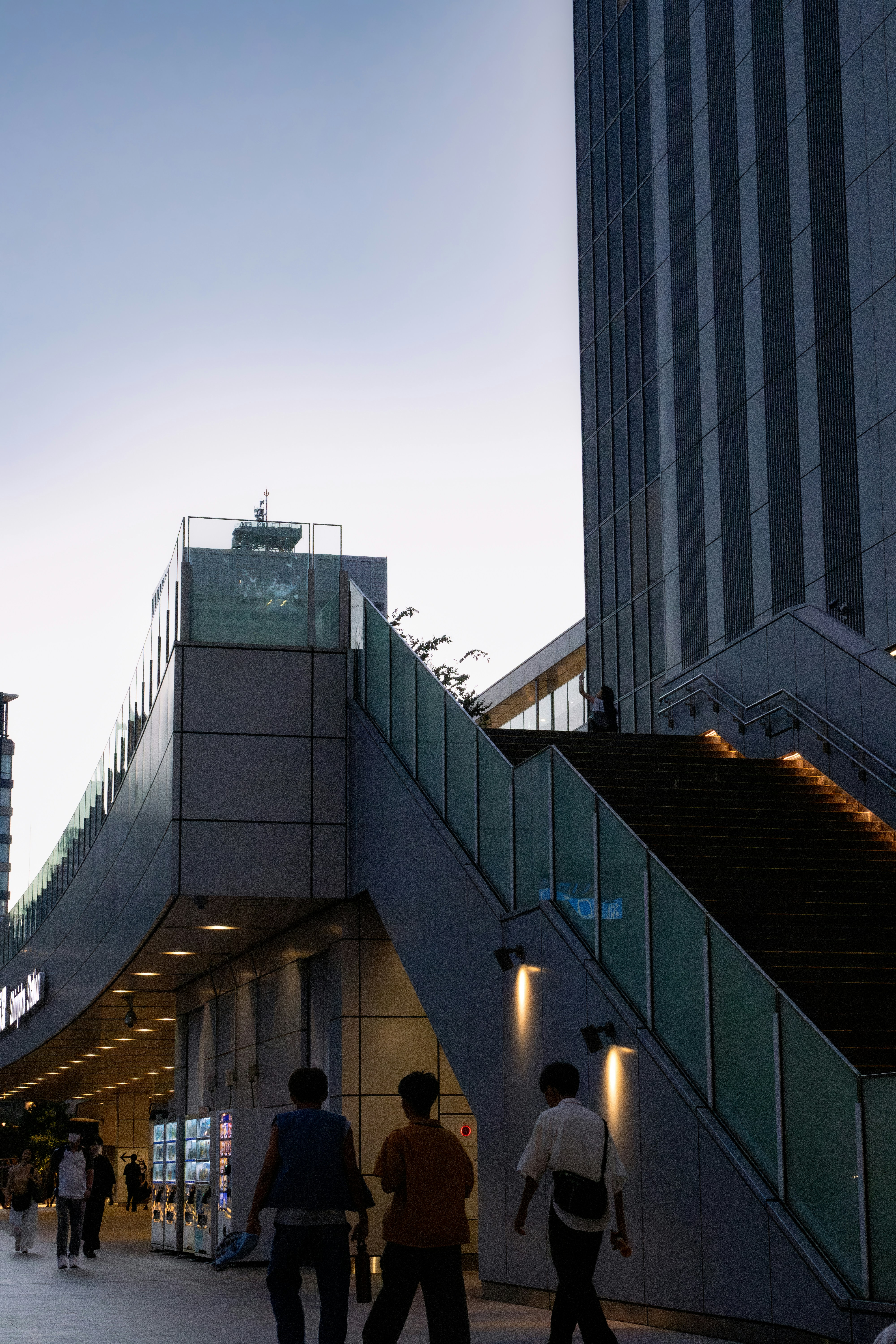 A view of Shinjuku Station in Tokyo, showcasing the modern architecture at dusk. The staircase with glass railings leads up to the station's upper level, with people walking through the area, capturing the bustling urban energy.