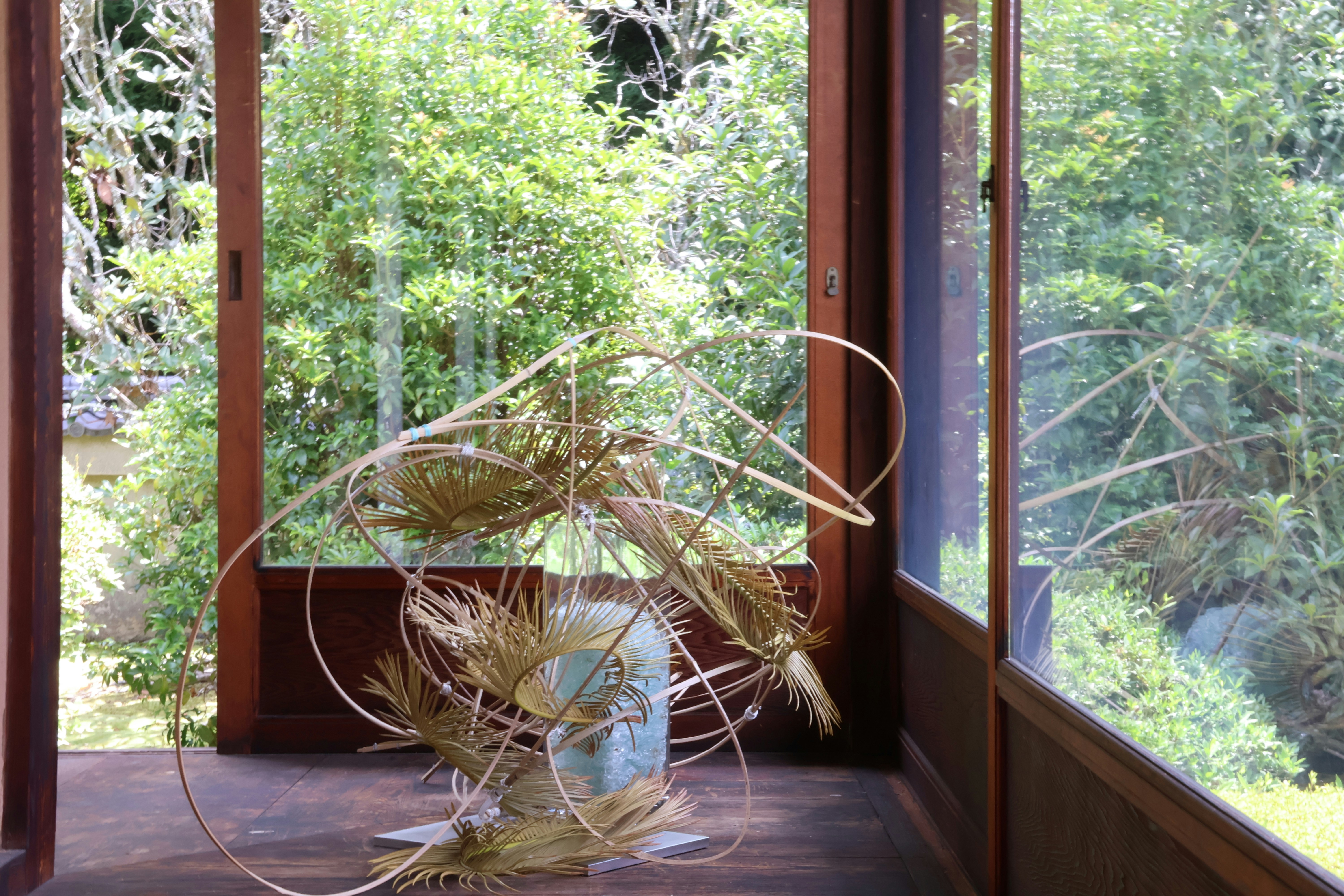 An elegant arrangement of dried palm fronds and wire art, set against a backdrop of lush greenery visible through glass doors.
