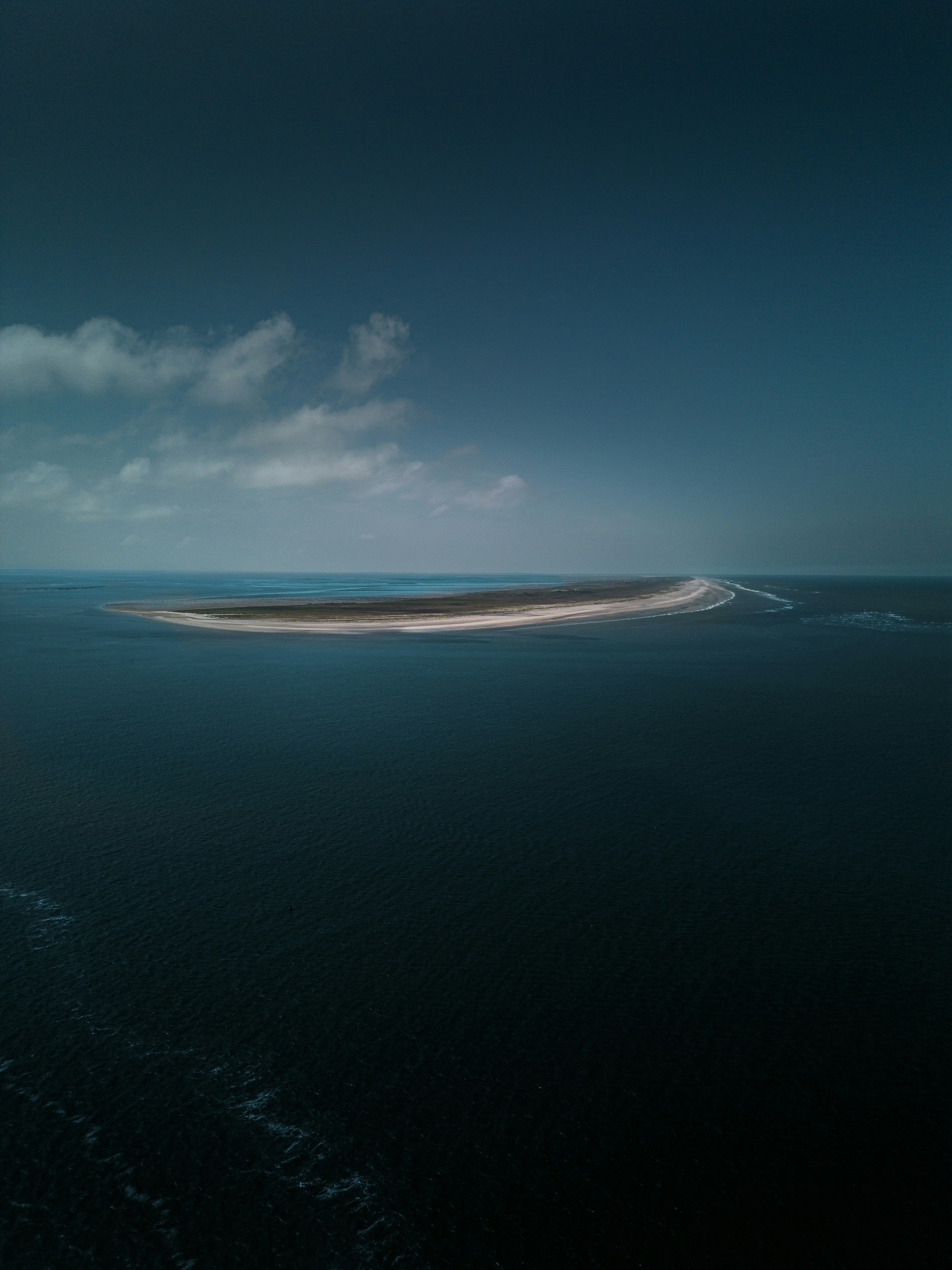 Aerial view of a crescent-shaped sandbar cutting through a calm deep-blue ocean under a muted sky.