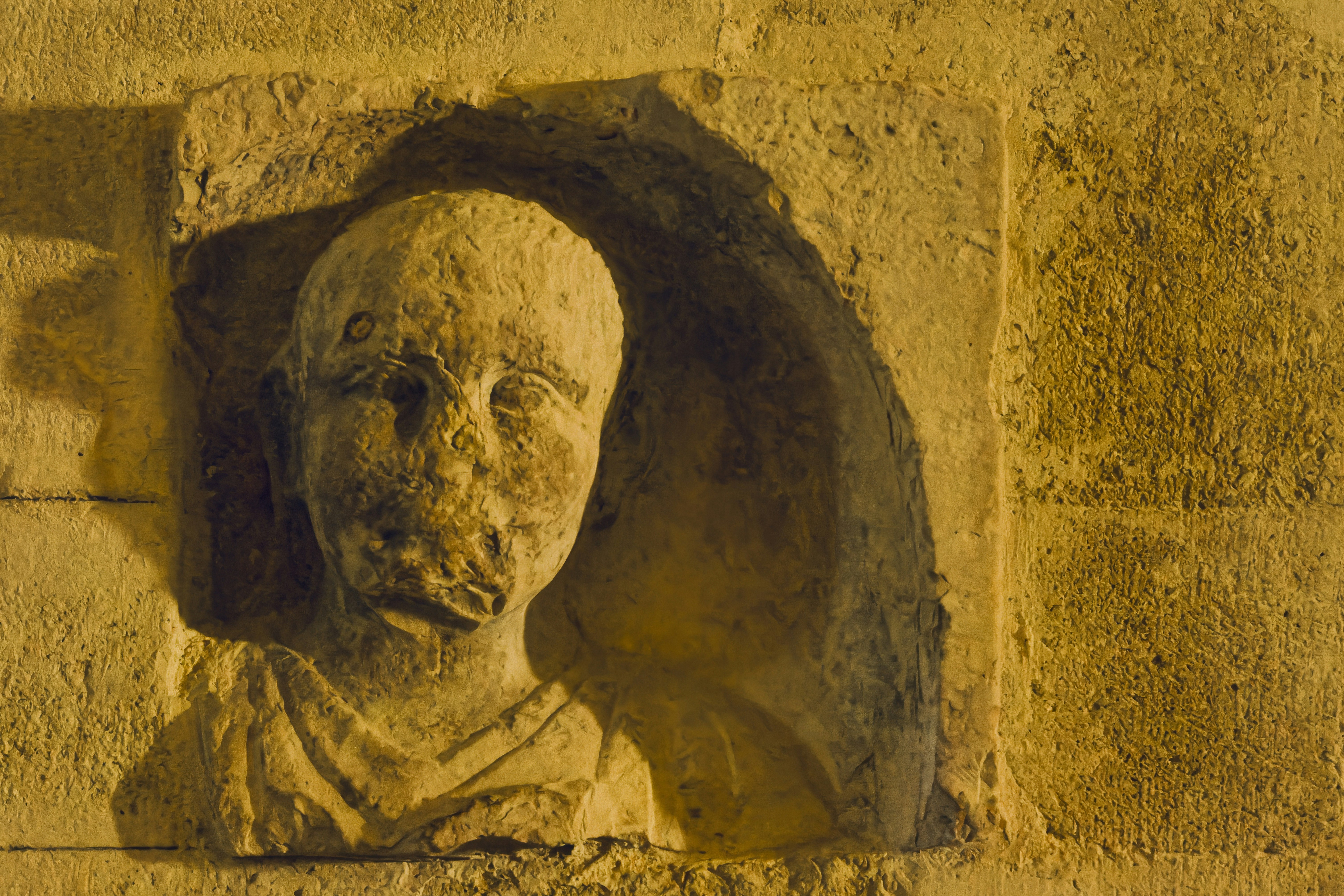 Ancient stone carving of a face displayed on a textured wall in a historical site during golden hour - Intricate stone carving of a human face is set against a textured wall in a historical site. The warm glow of golden hour highlights the features and details of the sculpture.