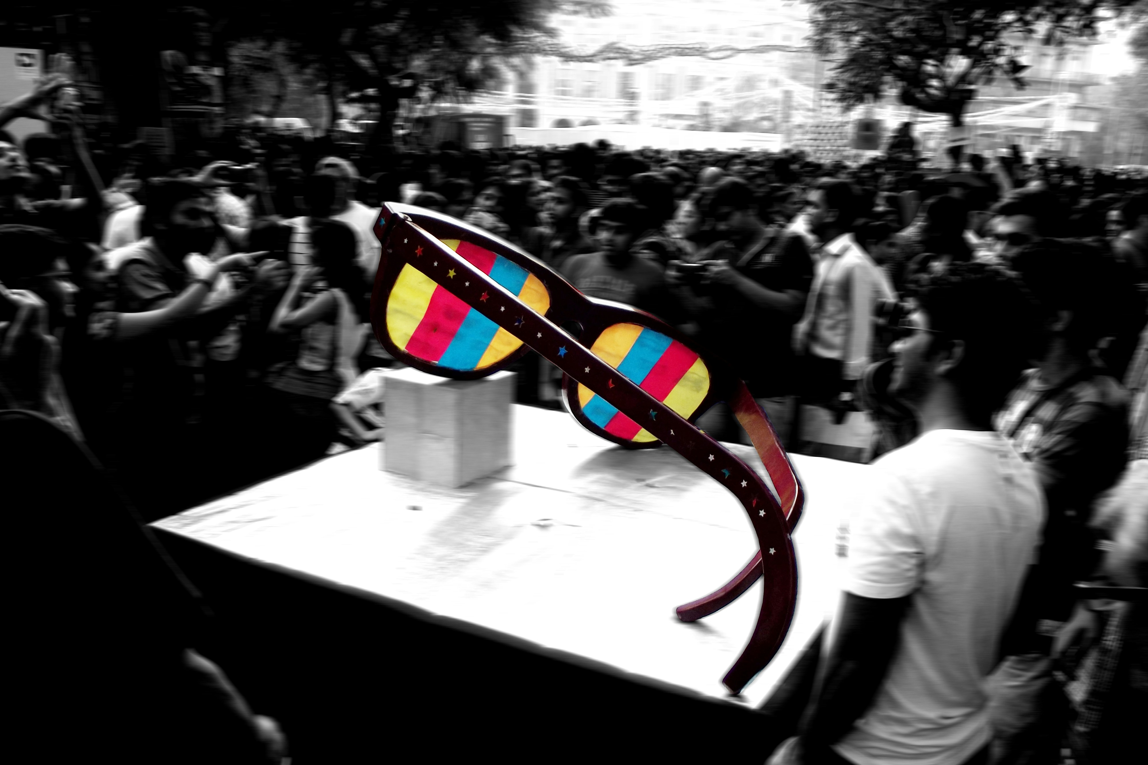 Colorful oversized sunglasses on a table surrounded by a black-and-white crowd.