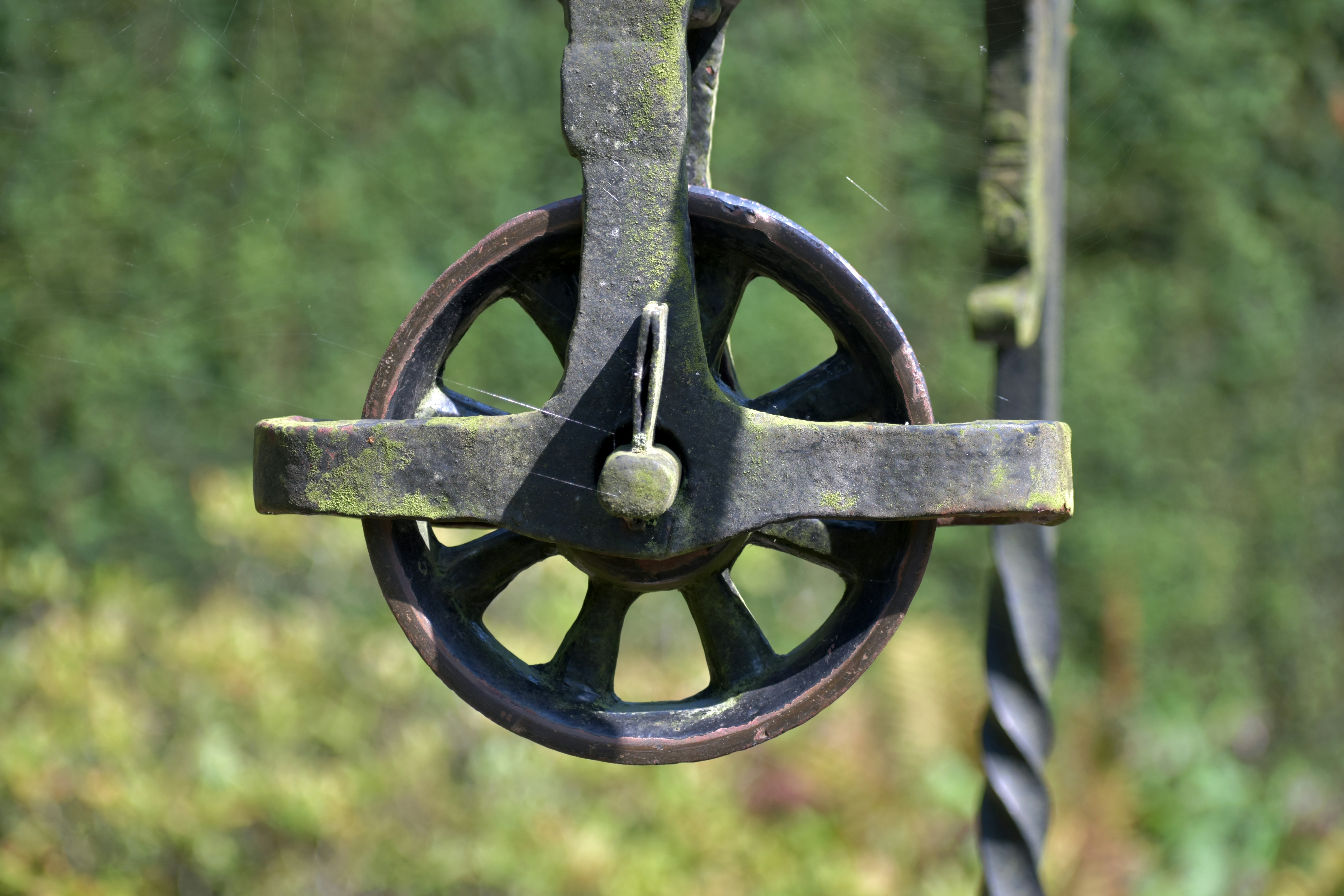 Close-up of a weathered pulley wheel hanging from a structure, showcasing intricate details and texture. The background features soft greenery, enhancing the historic feel.
